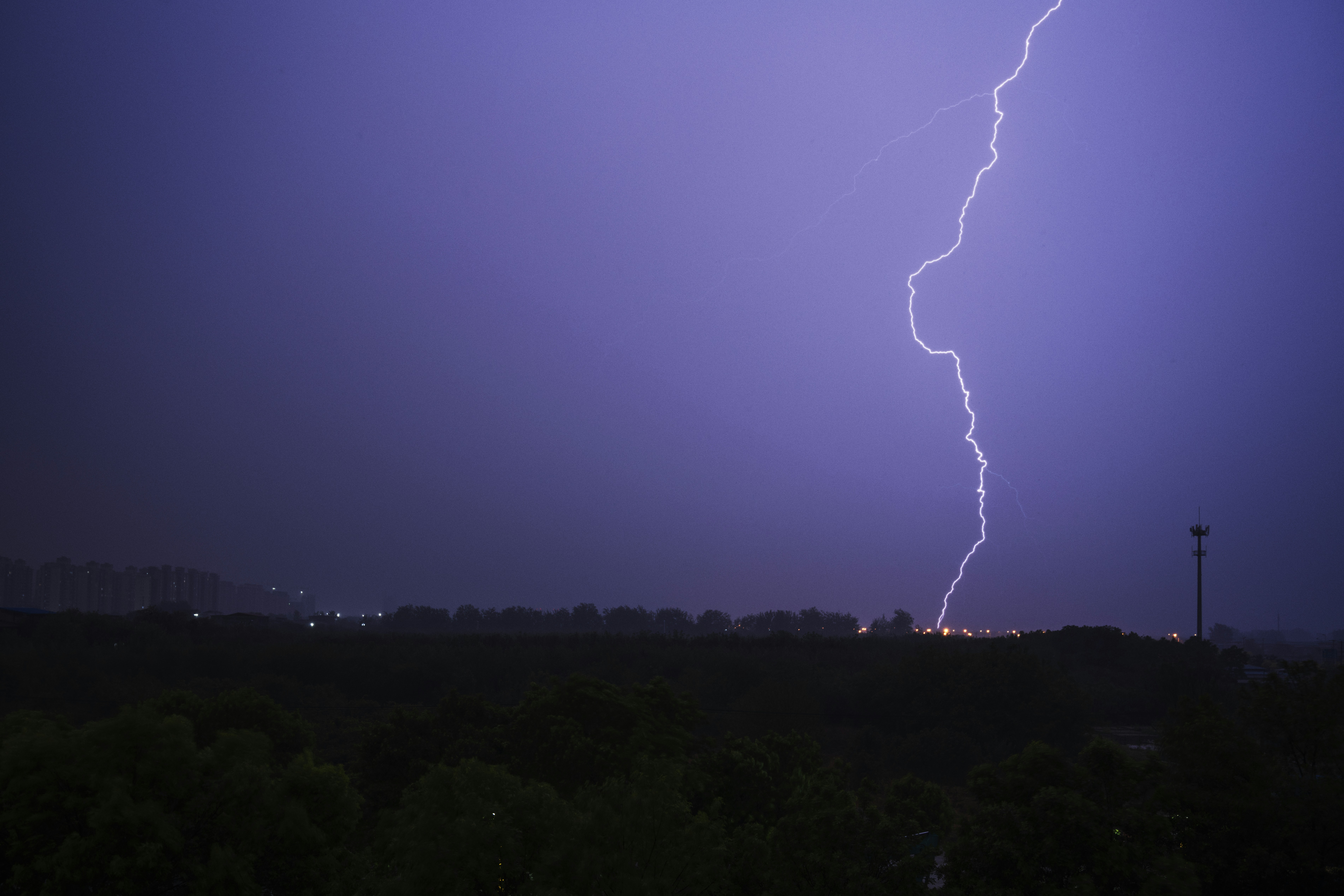 A single bright lightning bolt forks down across a dark, stormy sky above a flat landscape with distant city lights. The scene emphasizes raw electrical power against a calm ground silhouette; appears as a night-time, long-exposure capture.