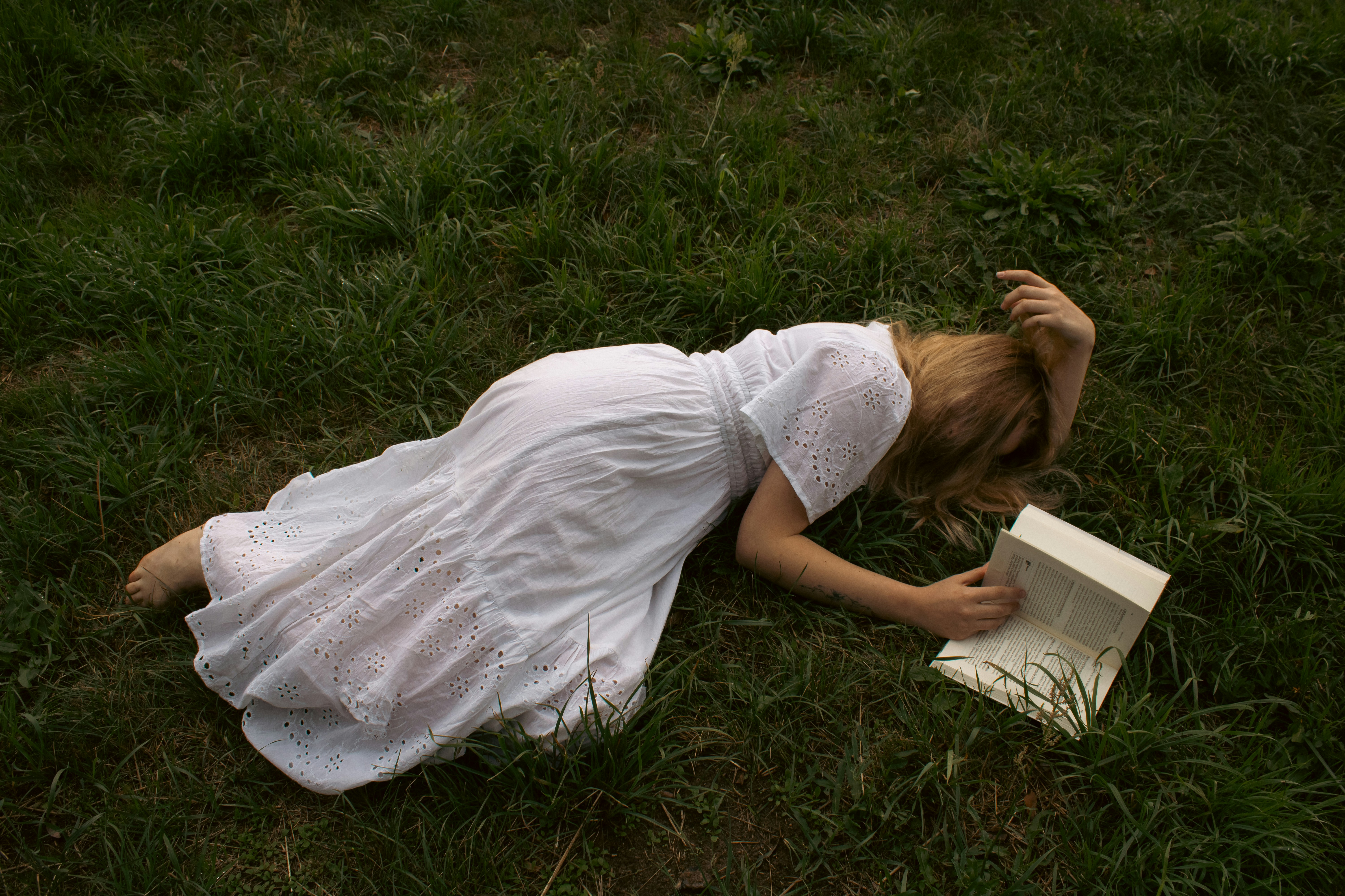 A girl in a white dress lies prone on grass with an open book beside her, creating a quiet, contemplative pastoral scene.