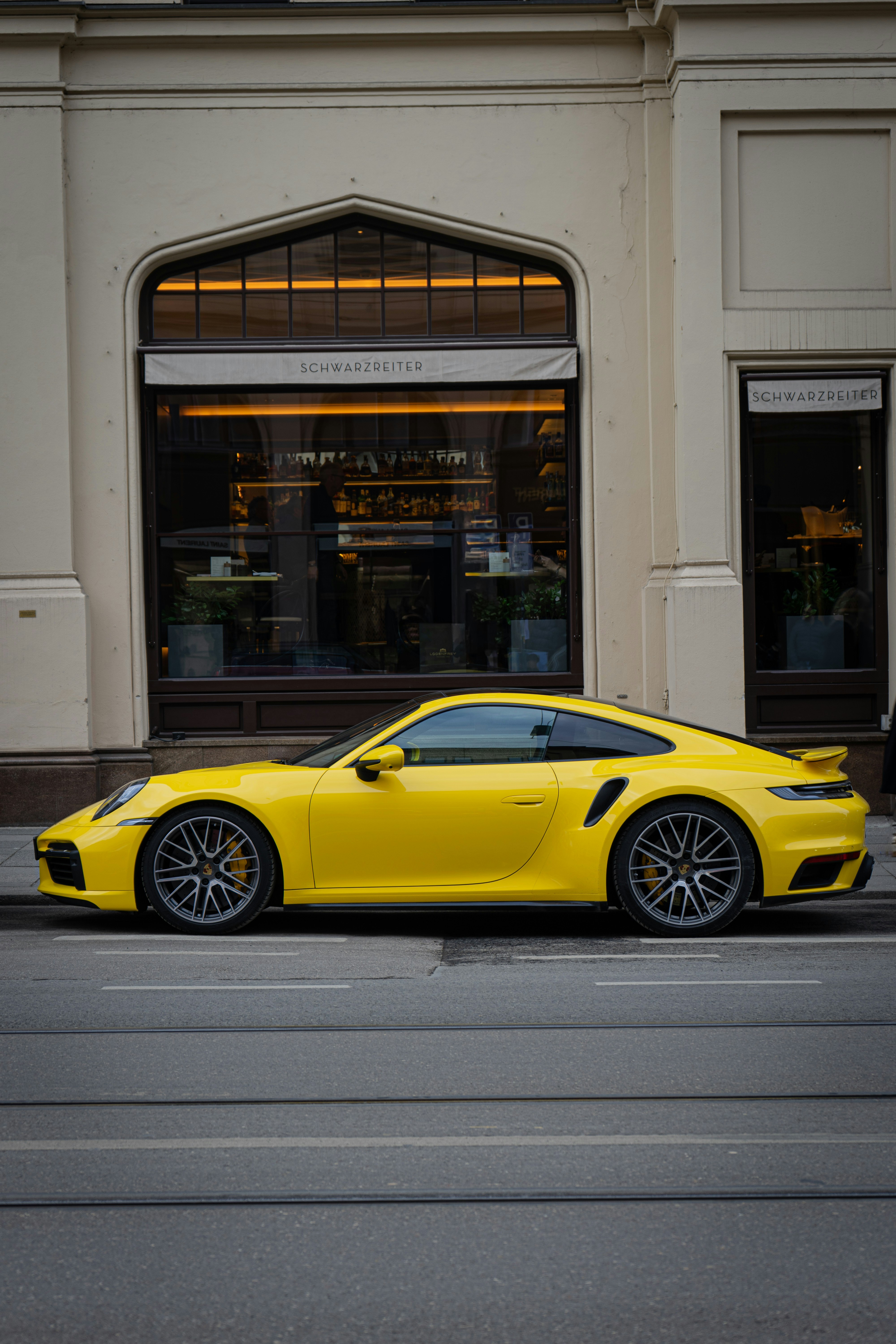 Bright yellow Porsche sits along a quiet city street with historic storefronts reflected in the windows behind it. The image captures a clean, urban scene with emphasis on the car’s silhouette and color. (Logo/ storefront reflections present).