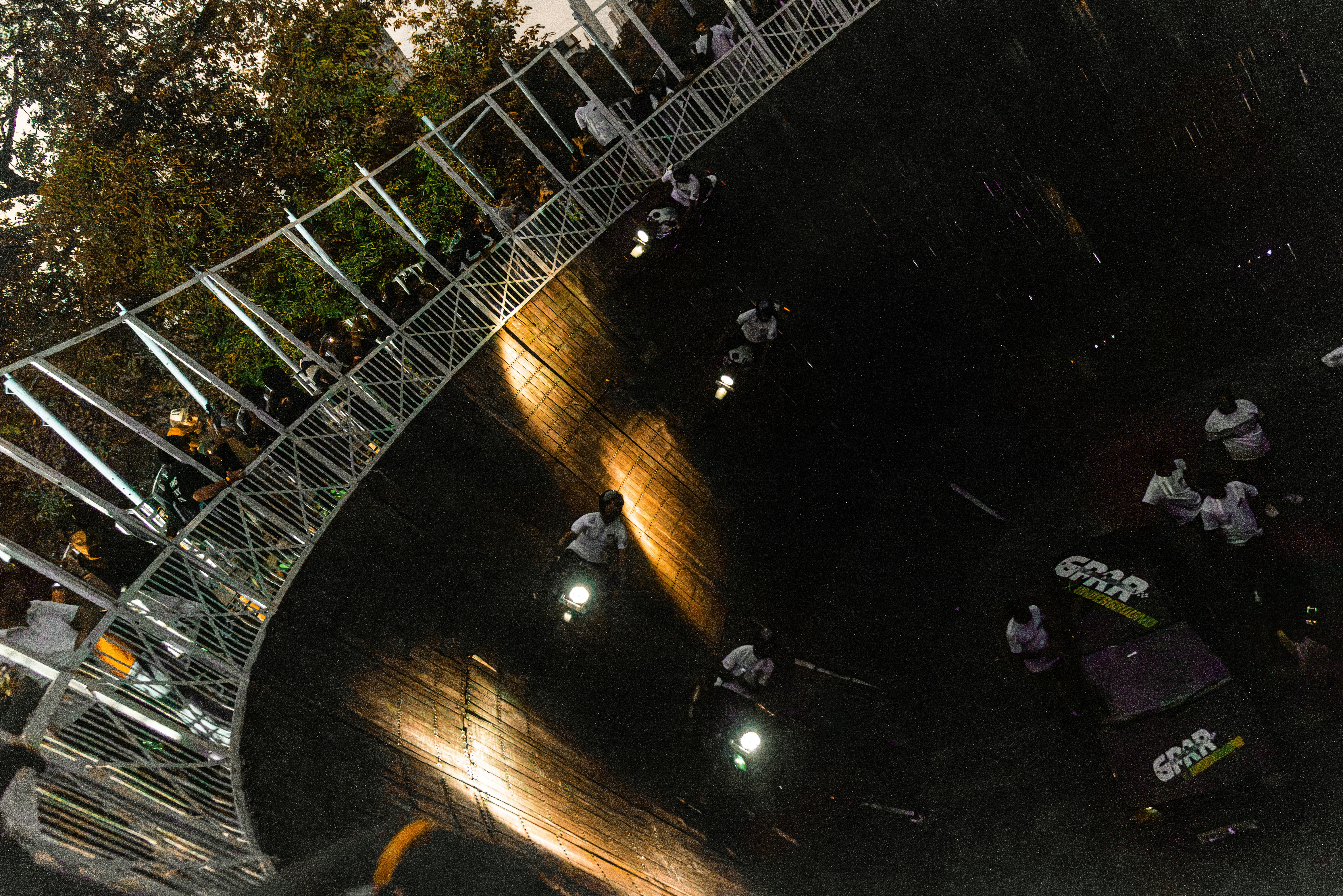 A dramatic view of the Maut Ka Kuan, or Well of Death, motorcycle stunt show at Richardson Cruddas in Byculla, Mumbai, India. Riders race motorcycles along the near vertical wooden walls of a circular well while spectators watch from the railing above. The photograph captures the danger, spectacle, and distinctive atmosphere of this traditional Indian carnival stunt performance.