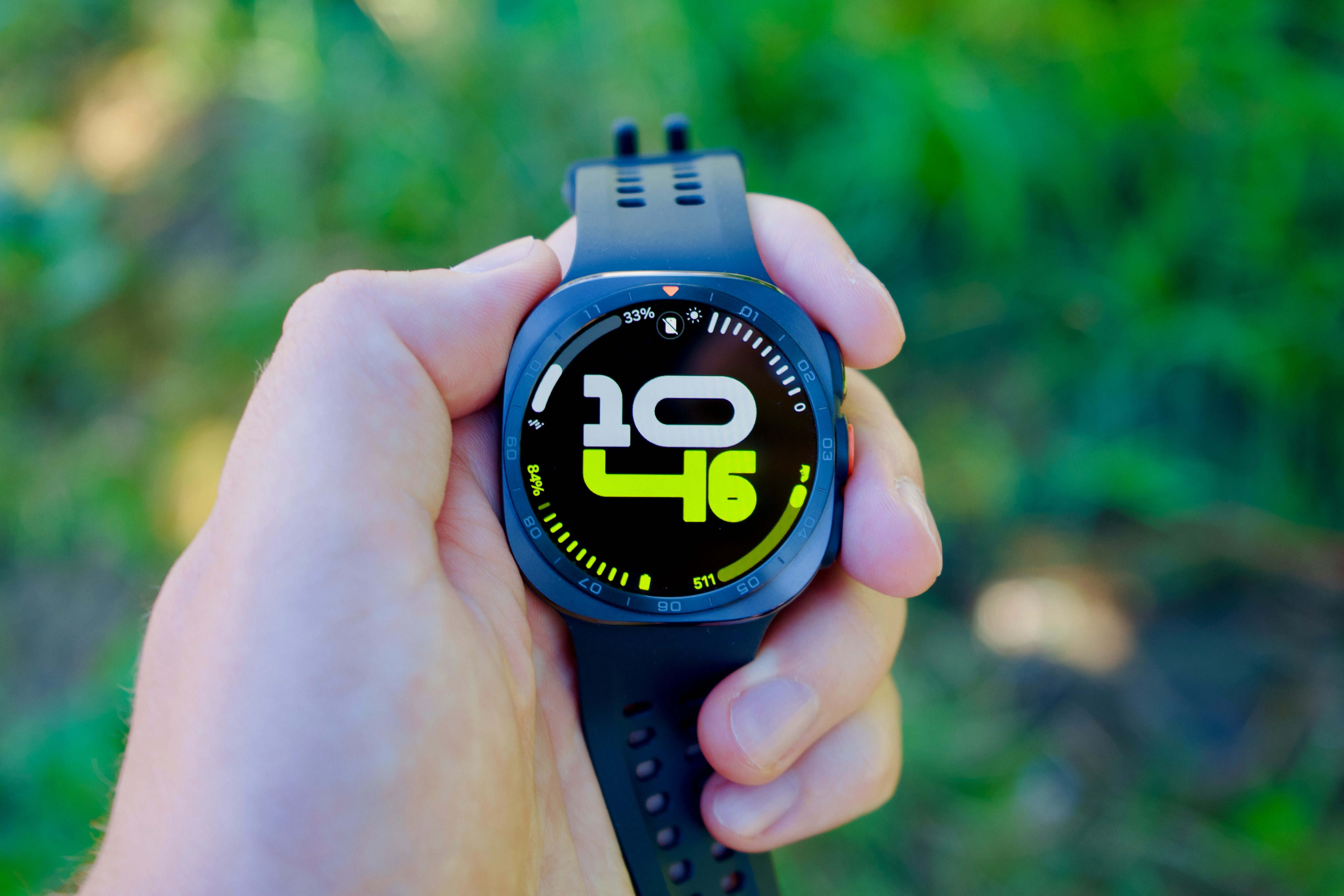 Bold smartwatch dominates foreground with a bright digital display; background blurred outdoors, emphasizing the device texture and strap.