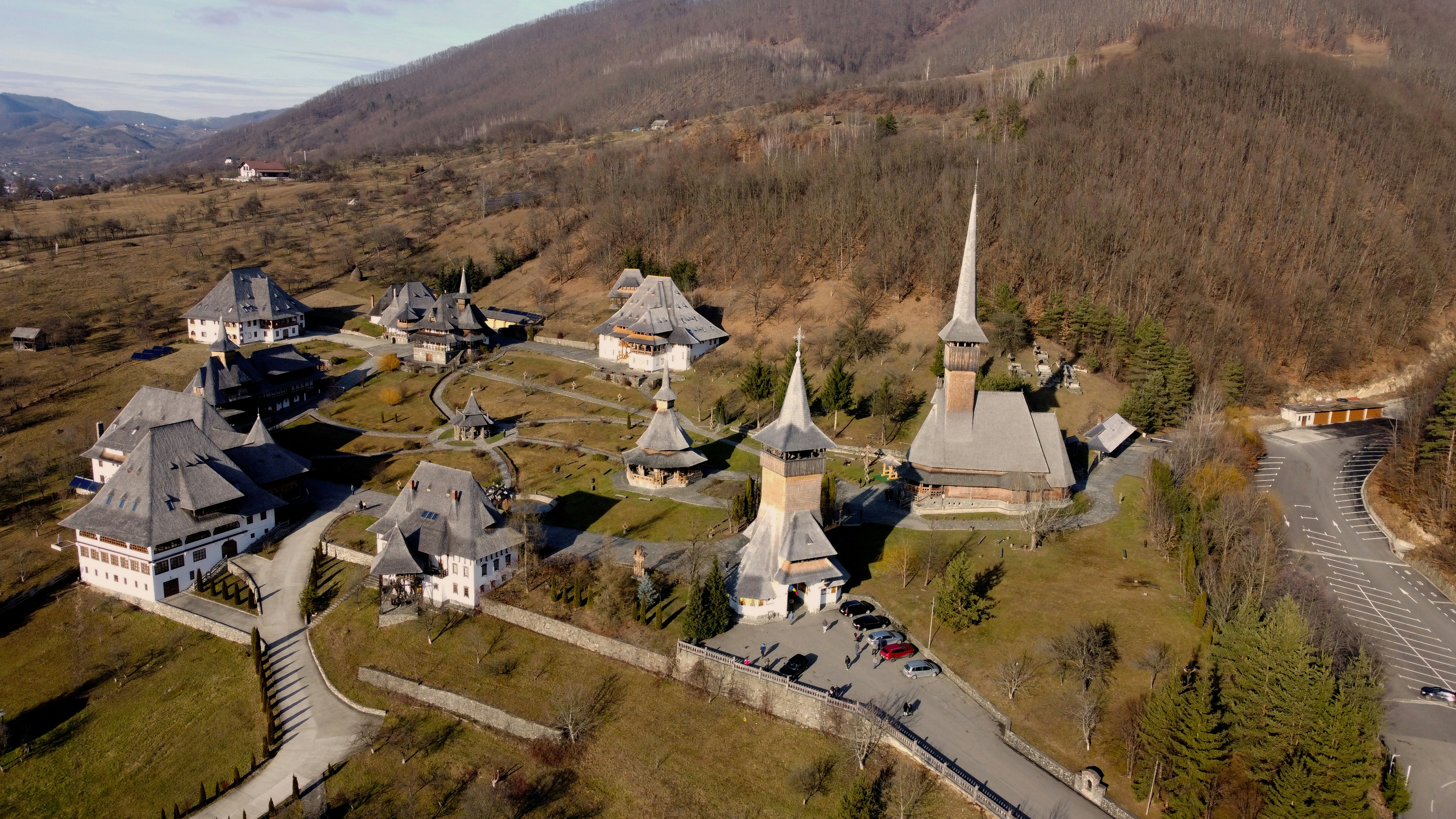 Aerial view of a hilltop complex of stone-and-wood structures with spired roofs set among trees and winding paths; drone-captured landscape highlighting architectural cluster and surrounding forested hills.