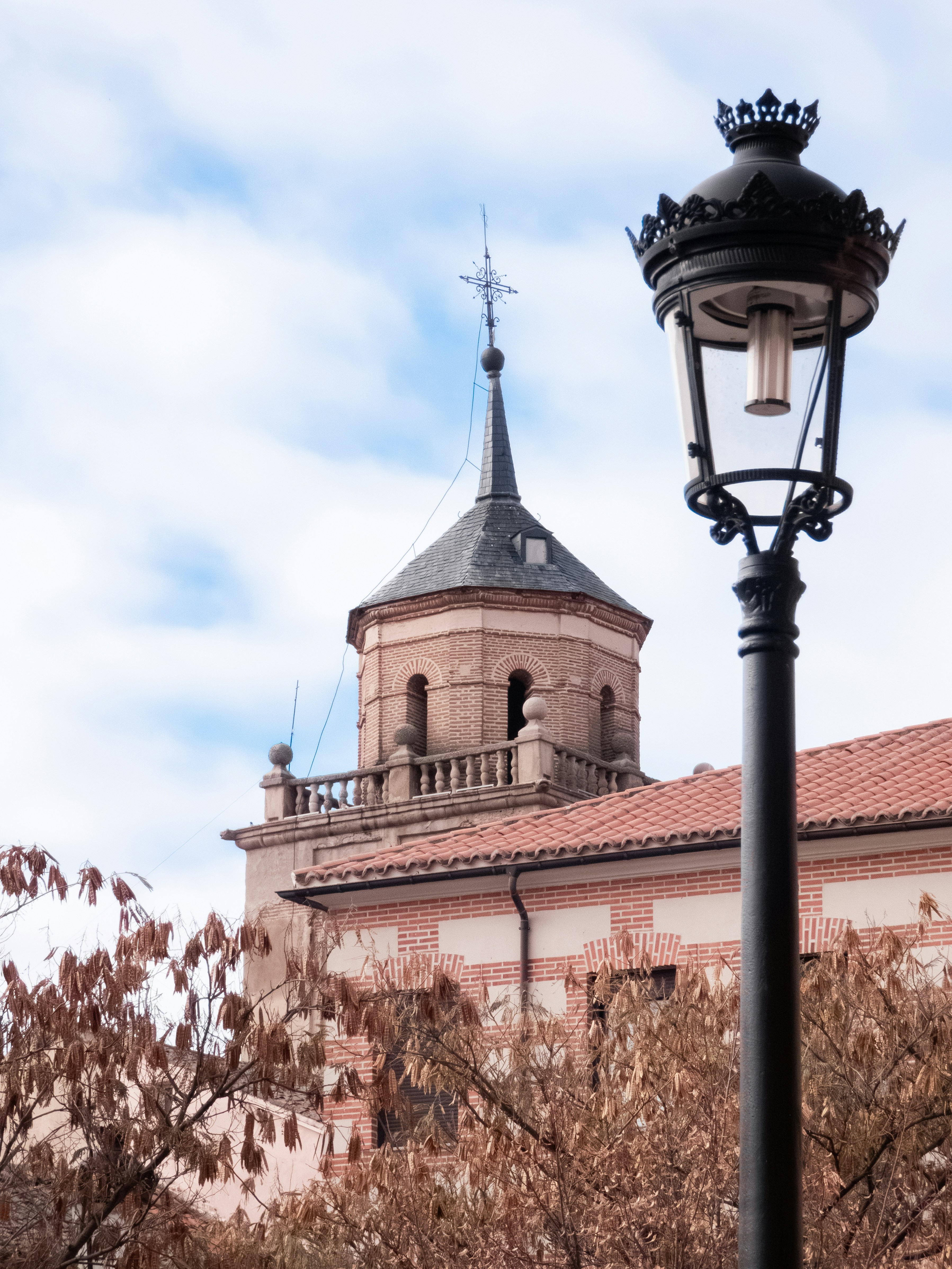 A low-angle shot depicting a black wrought-iron street lamp post on the right foreground. In the background stands a brick church structure with an octagonal tower topped by a slate roof spire and iron cross. Dry, leafless tree branches occupy the lower-left foreground against a blue sky with scattered white clouds.