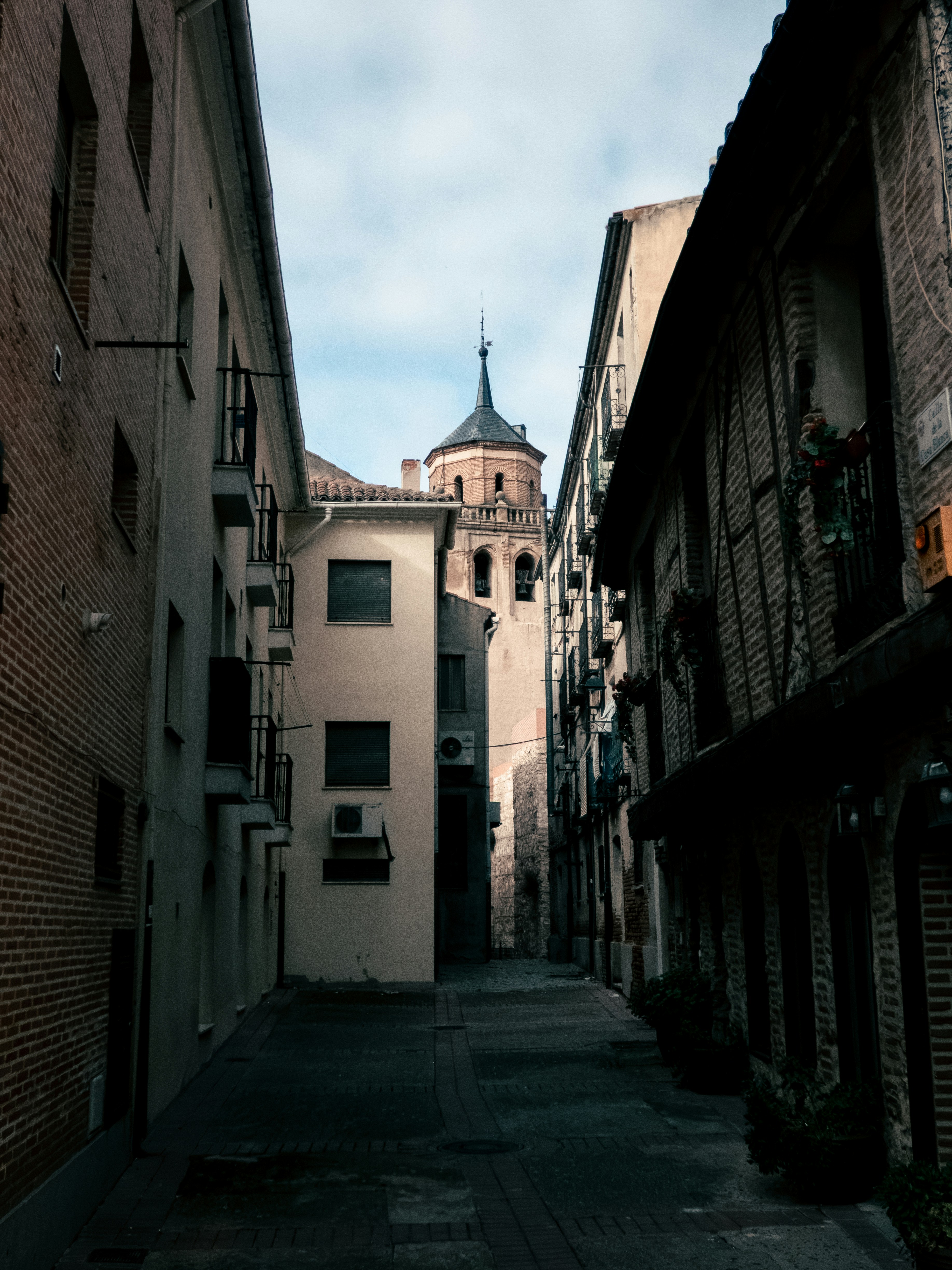 A narrow paved alleyway bordered by multi-story brick and stucco buildings on both sides creating a strong perspective line. In the distance, at the end of the corridor formed by the buildings, an octagonal brick church tower with a spire is visible under an overcast sky.