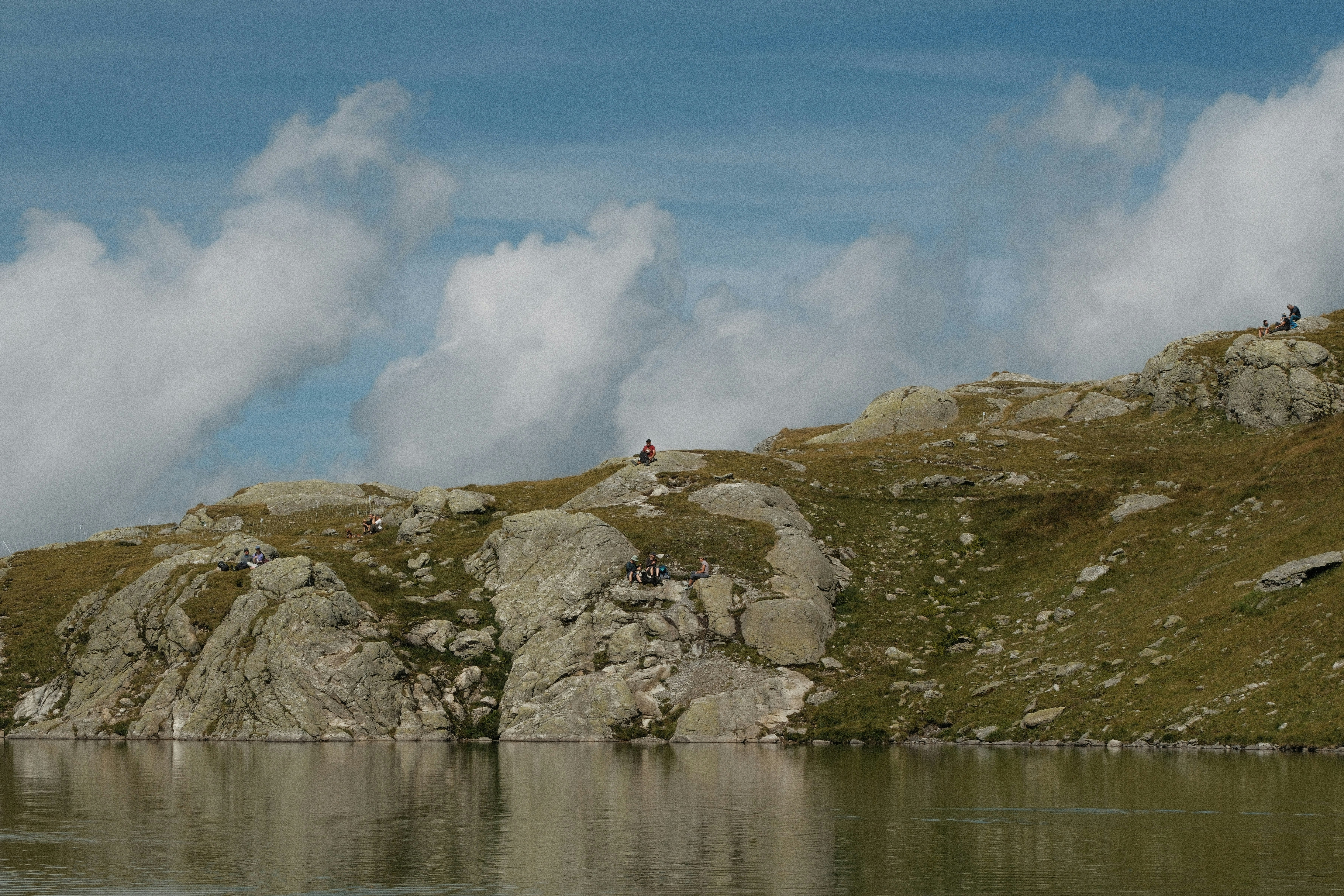 Climbers traverse rocky ridge above a calm lake with a clear sky and scattered clouds; a small group is visible amidst rugged terrain. The scene emphasizes scale and the quiet drama of highland exploration.