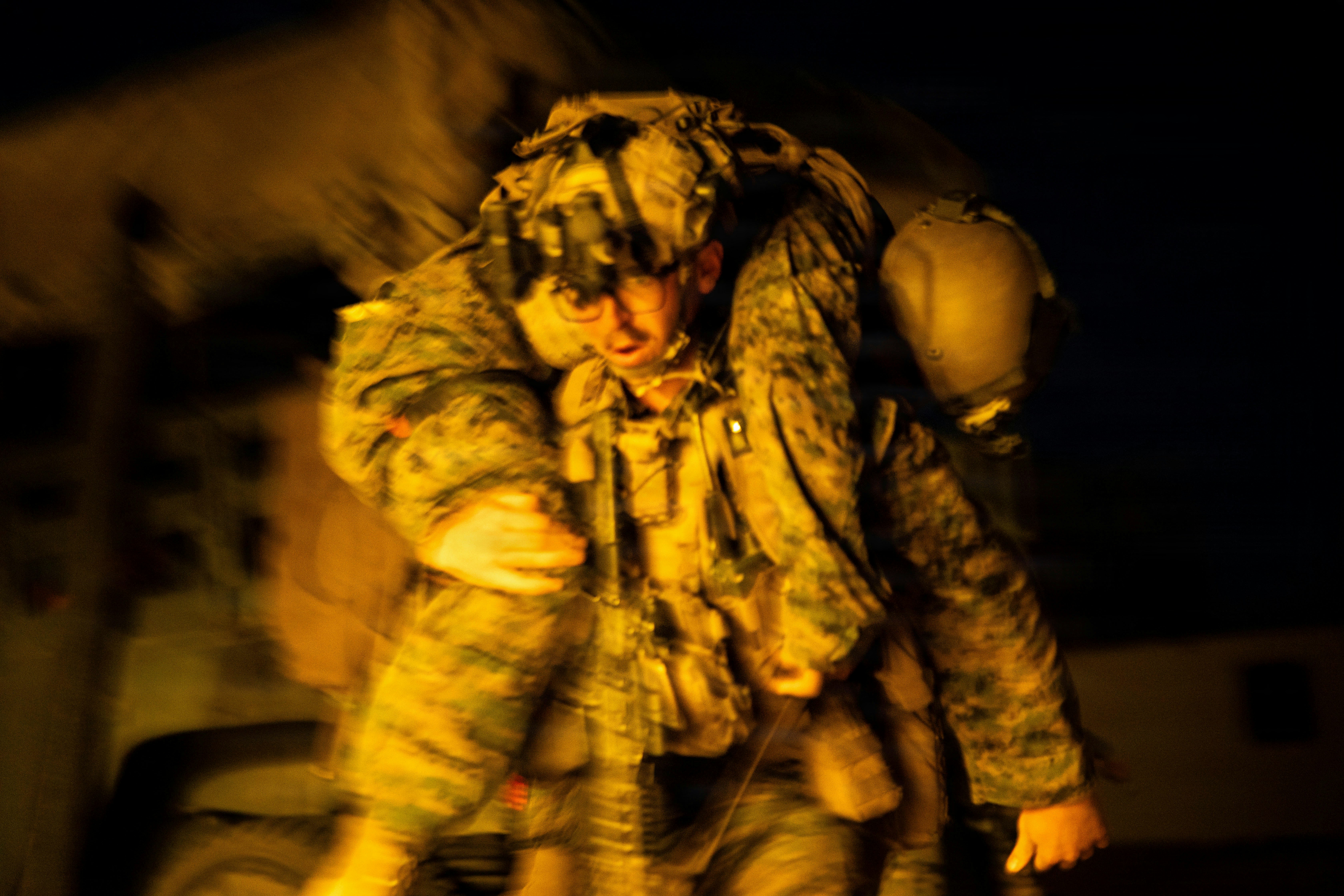 Soldier helps a comrade during a nighttime operation, both in camouflage gear and moving through dim lighting.