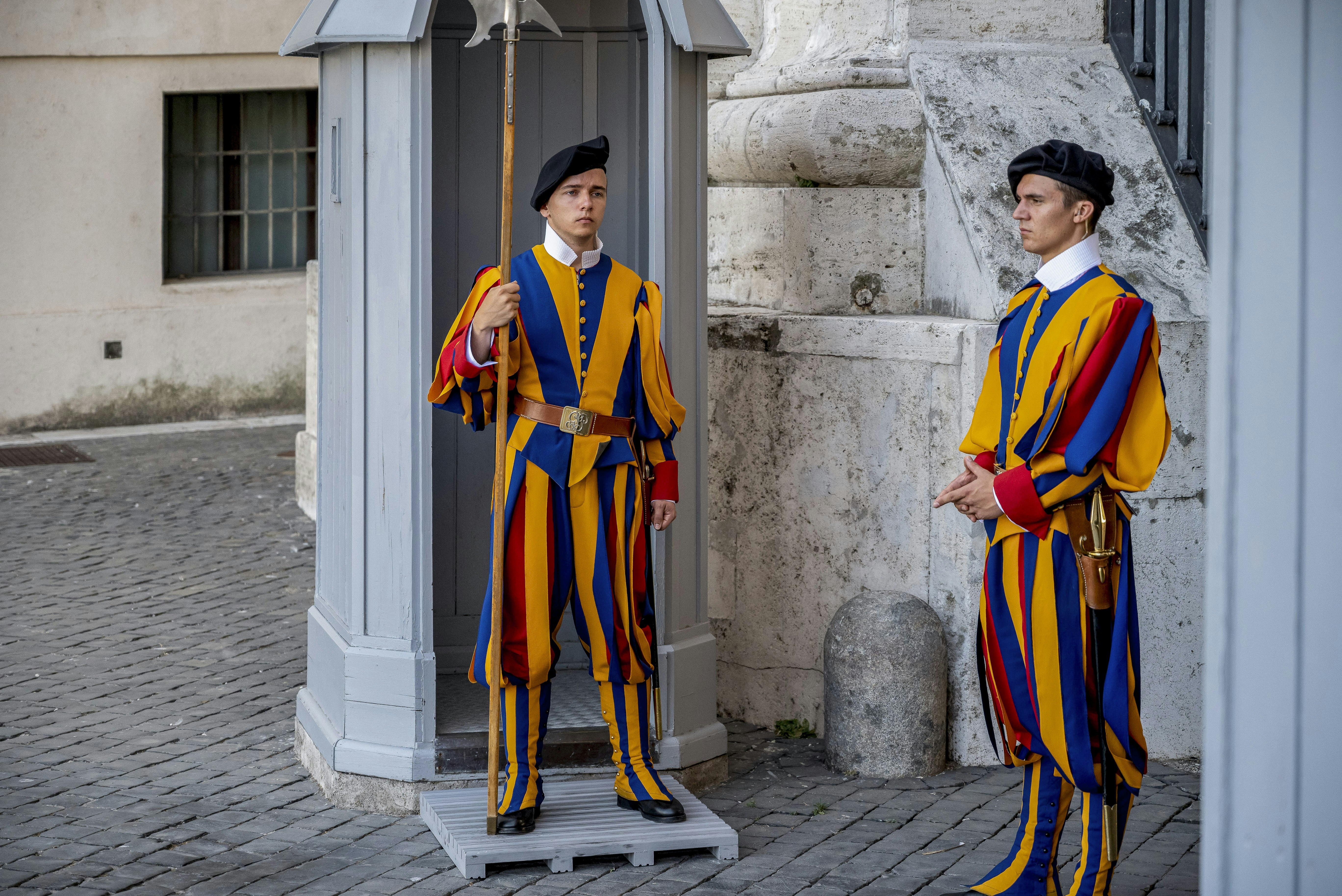 Two Swiss Guards stand in vibrant striped uniforms near a historical doorway, one inside a small guard booth and the other leaning against a column, cobblestone ground beneath them.