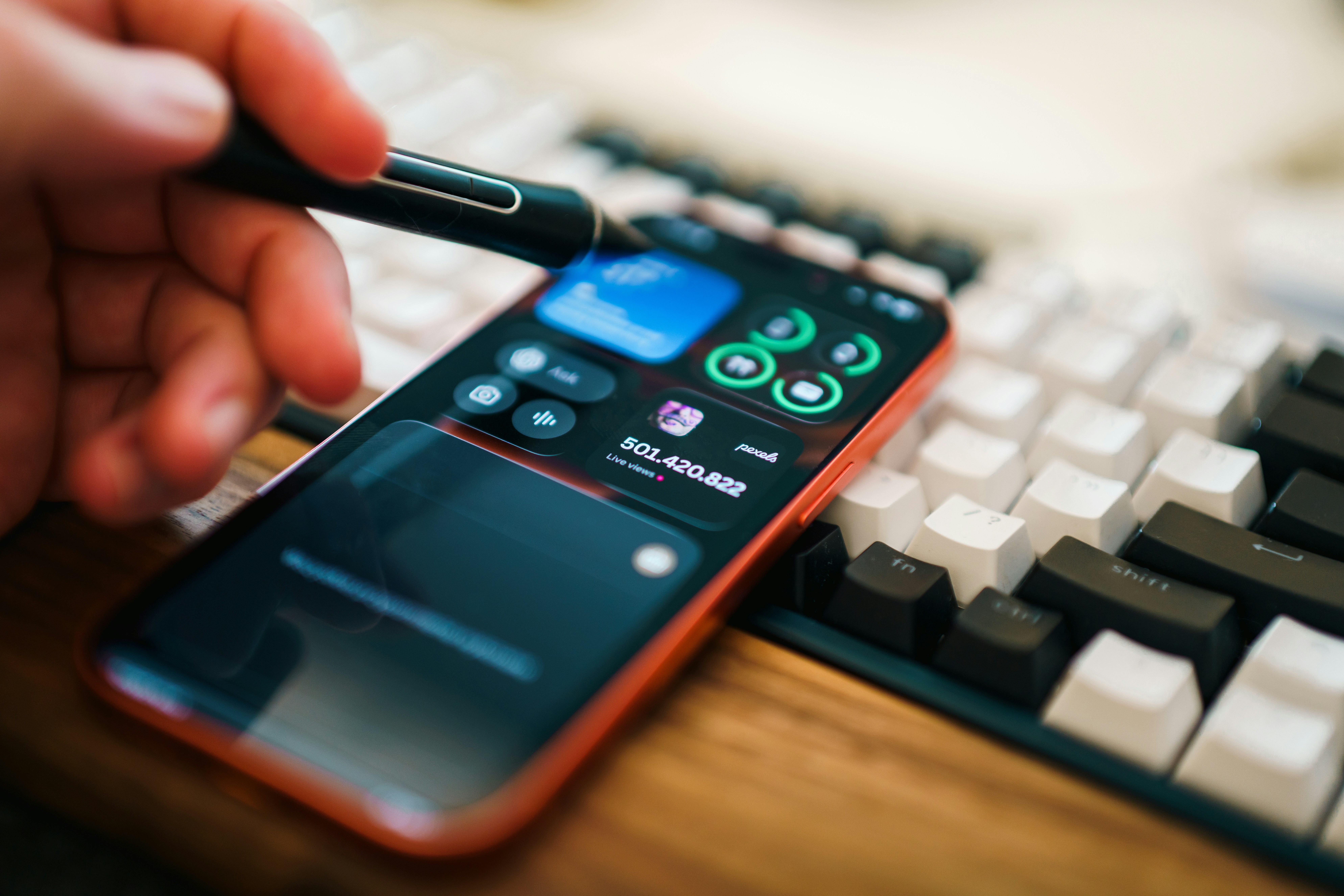 A close-up of a hand holding a stylus over a smartphone on a mechanical keyboard, highlighting digital input within a tactile desk setup.