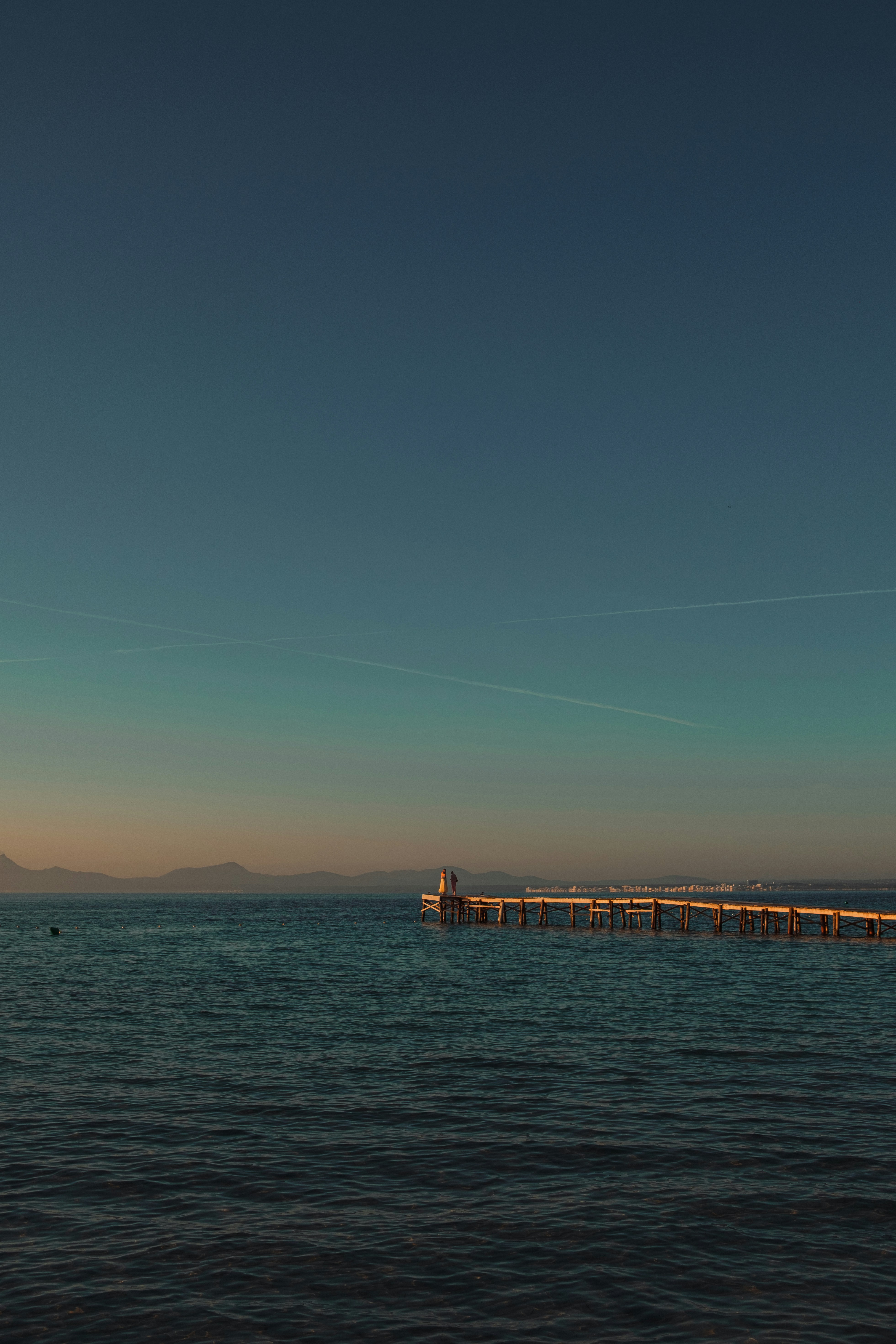 A wide minimalist vertical composition featuring two distant figures standing at the tip of a long wooden pier in Mallorca. Rich gradient tones transition from a deep teal sky to a warm golden sunrise horizon over calm waters, creating a serene sense of scale and isolation.
