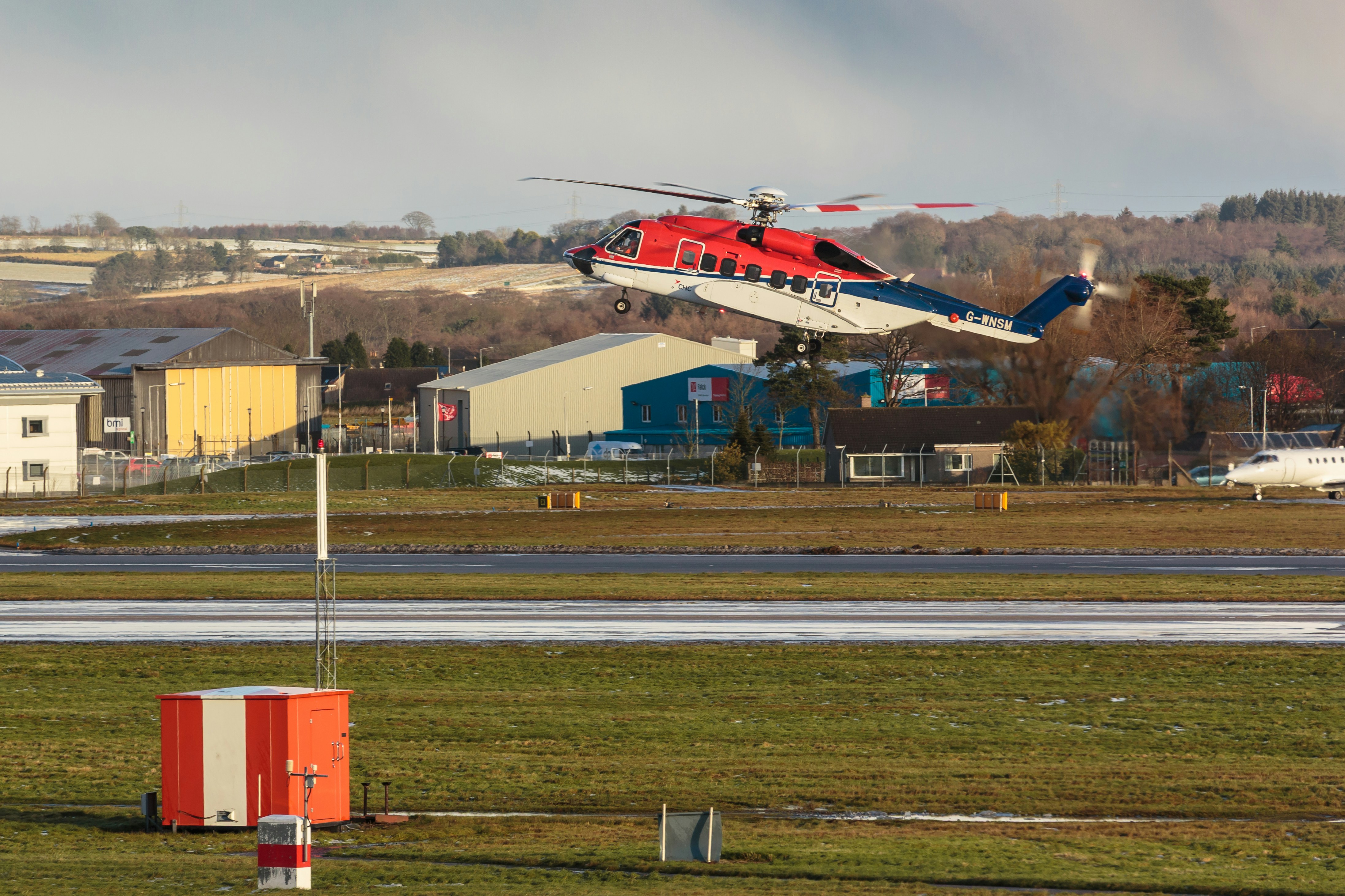 A red-and-white helicopter rises off a grassy airfield, with hangars and distant trees in the background under a clear sky.