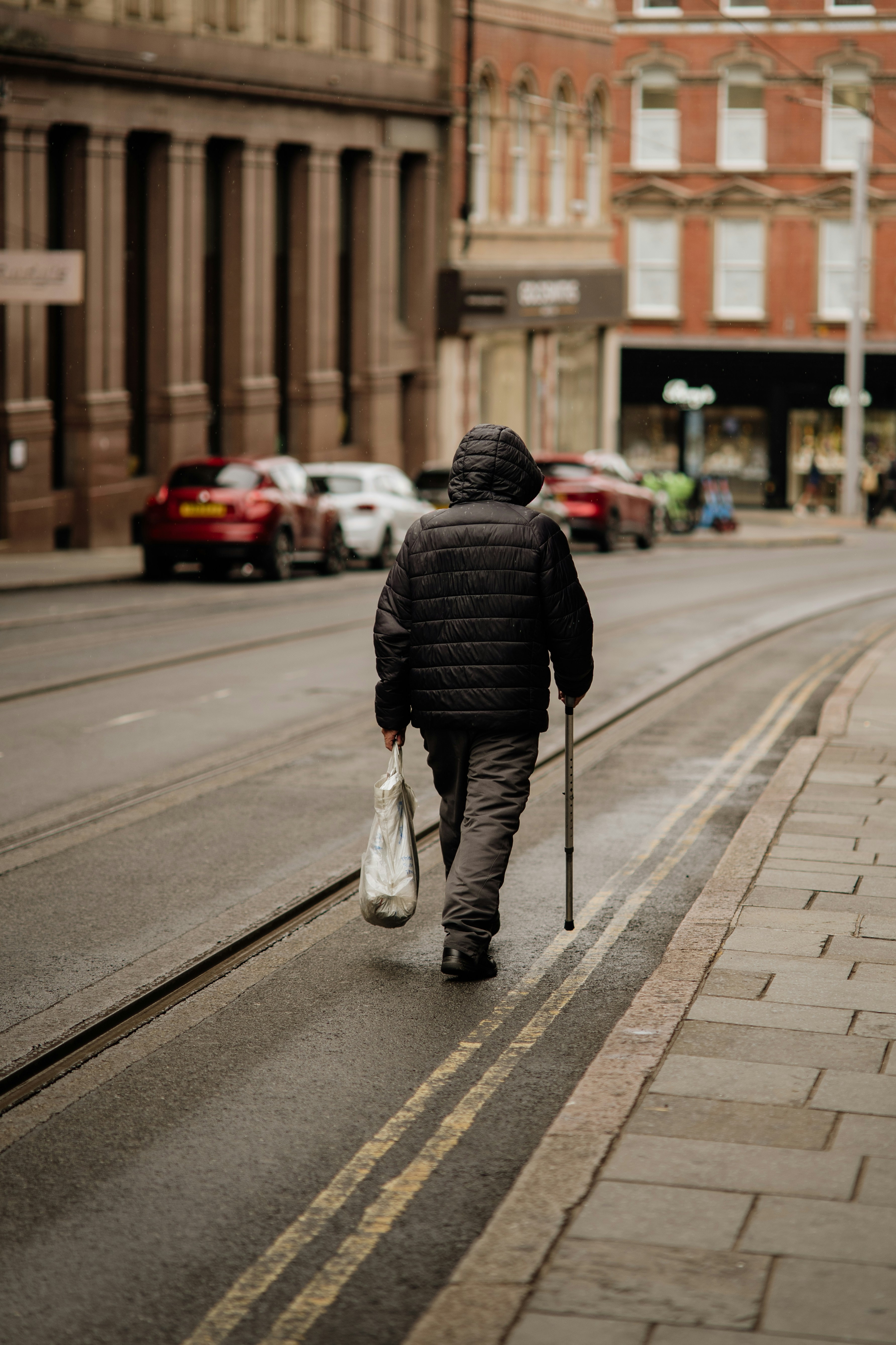 Man with a Hood and Walking Cane Crossing a Cobblestone Street
