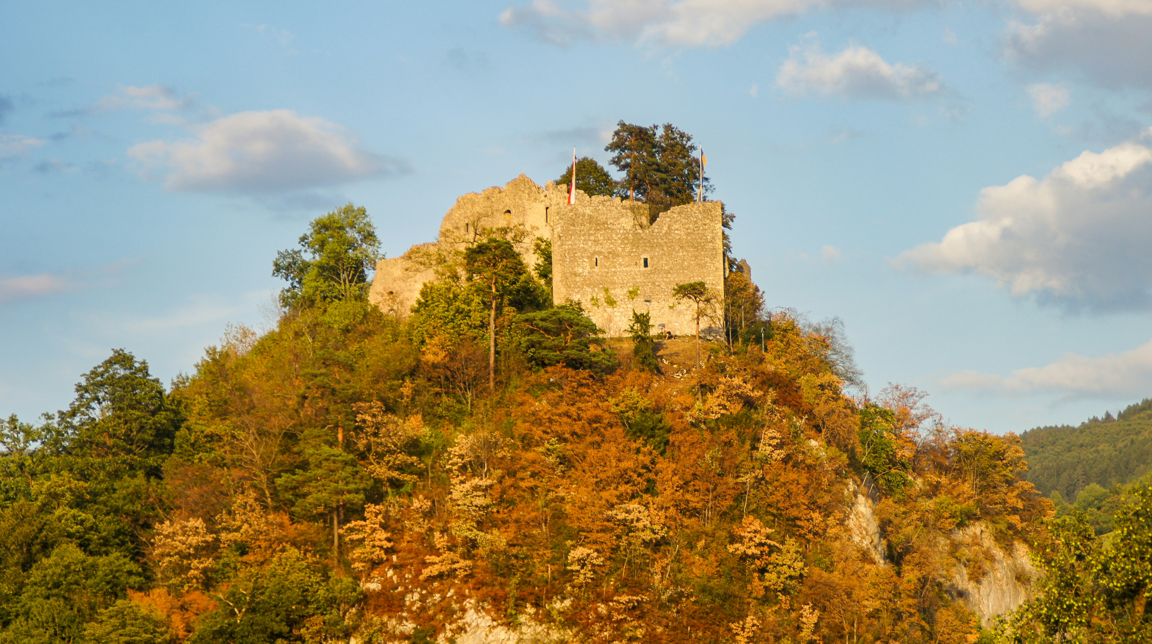Autumnal ruin perched on a rocky hill with a forested slope in warm hues under a blue sky with clouds.