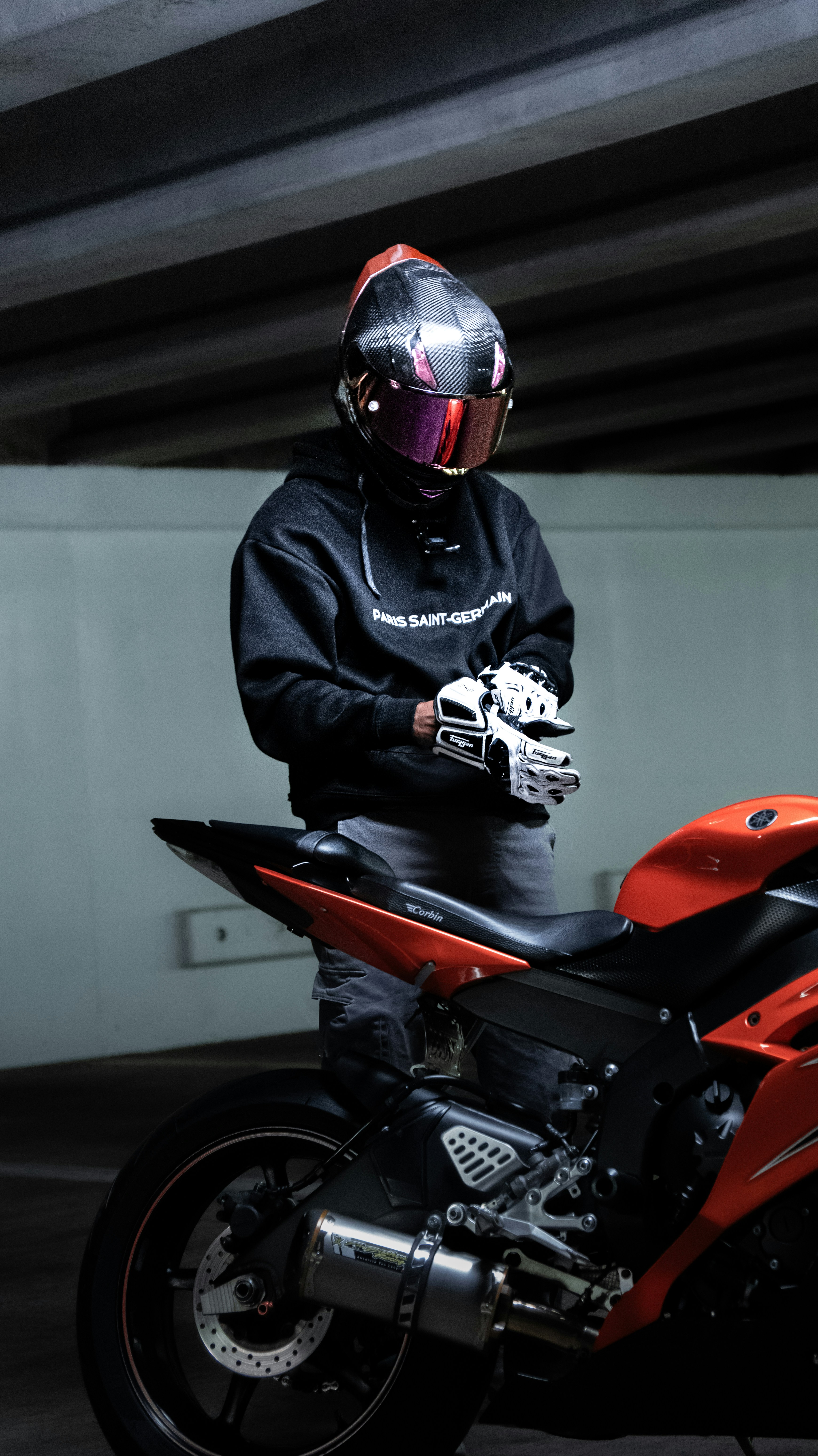 Rider in black hoodie and carbon-fiber helmet stands beside a red sport motorcycle in an industrial underpass, hands resting on gloves, moody lighting and sharp contrast.