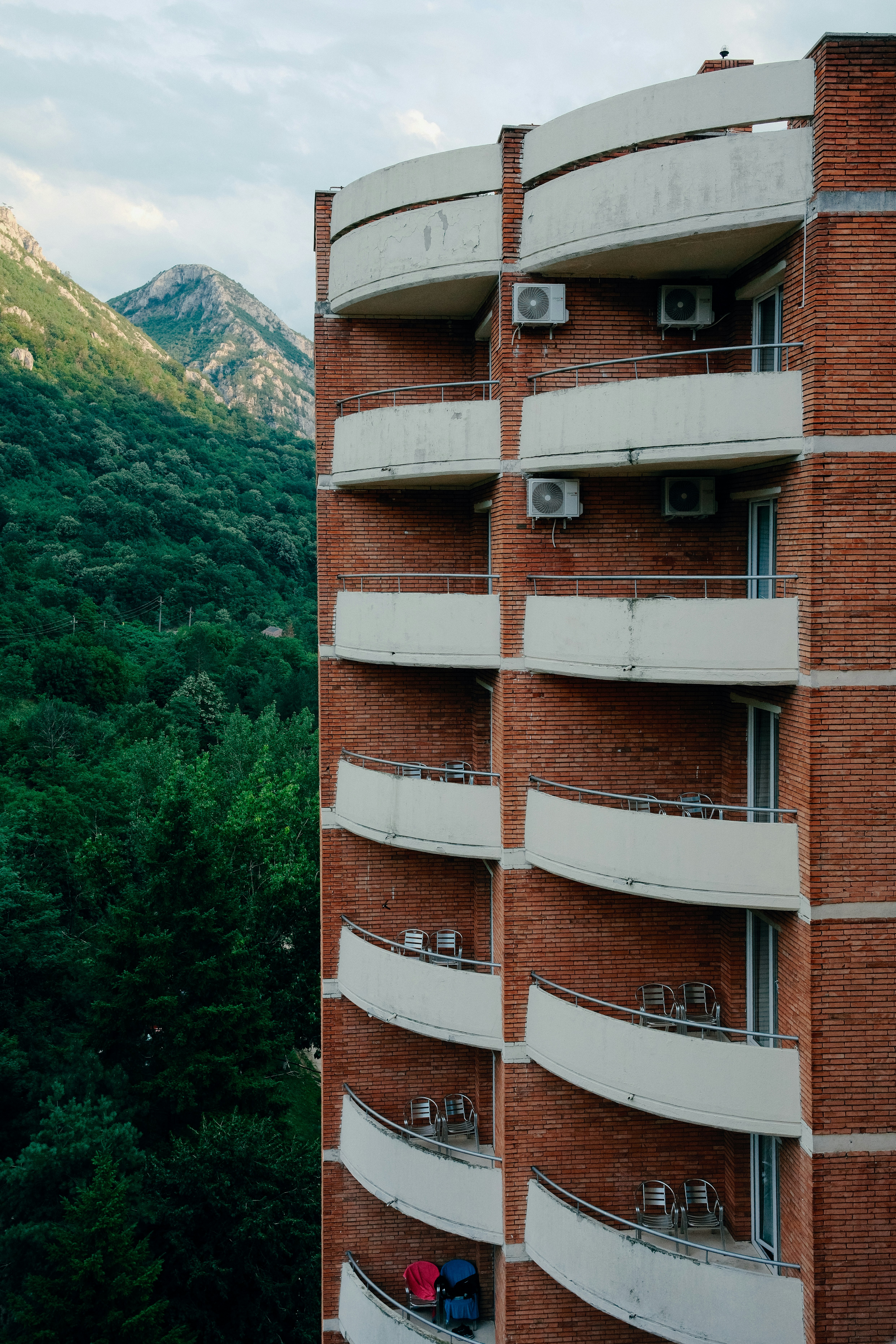 A red brick apartment building with curved balconies overlooking a forested mountain slope
