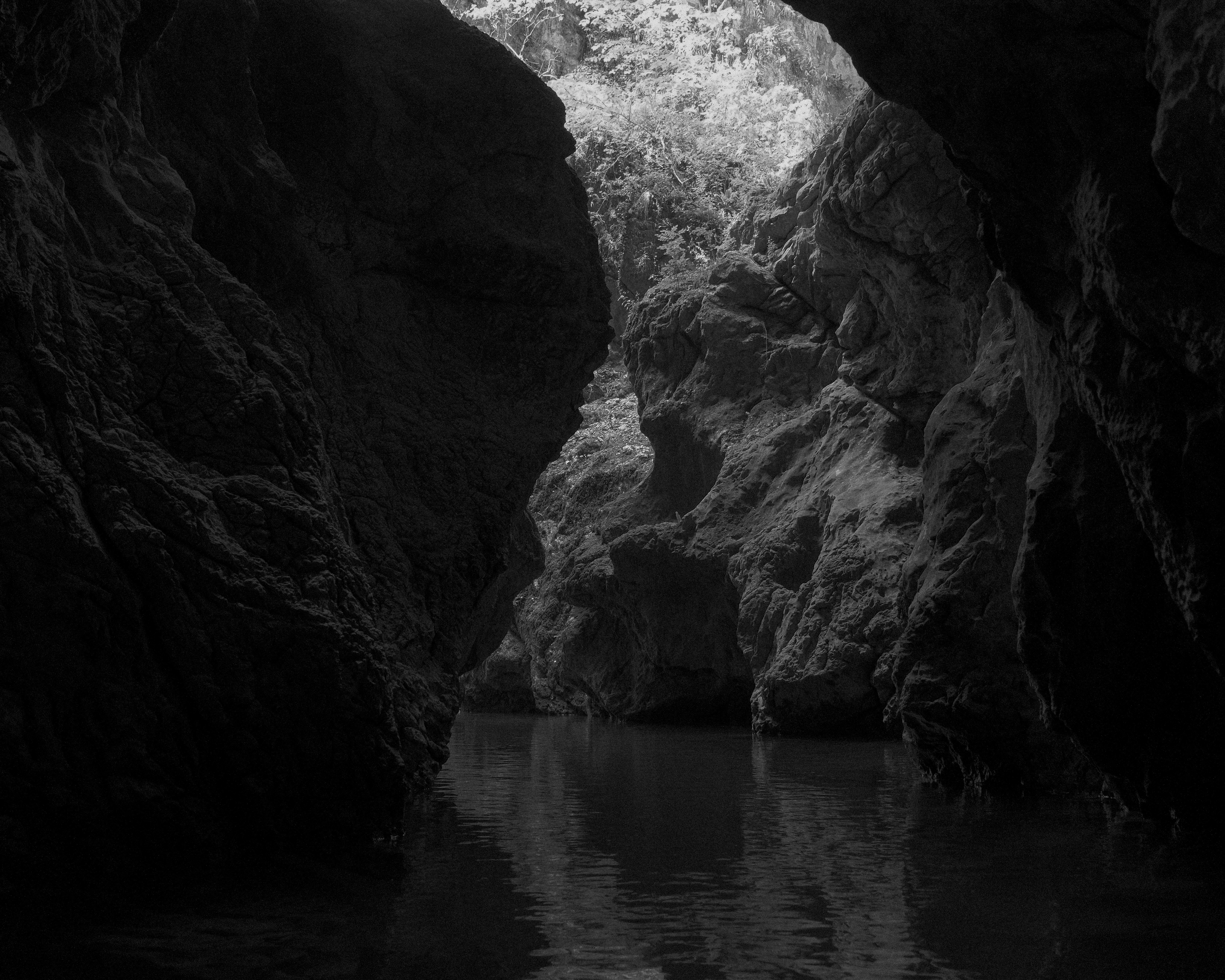 A monochrome scene of a narrow canyon with calm water reflecting carved rock walls, inviting the viewer into a quiet crevice between towering formations. A logo-free, landscape capture emphasizing texture and contrast.