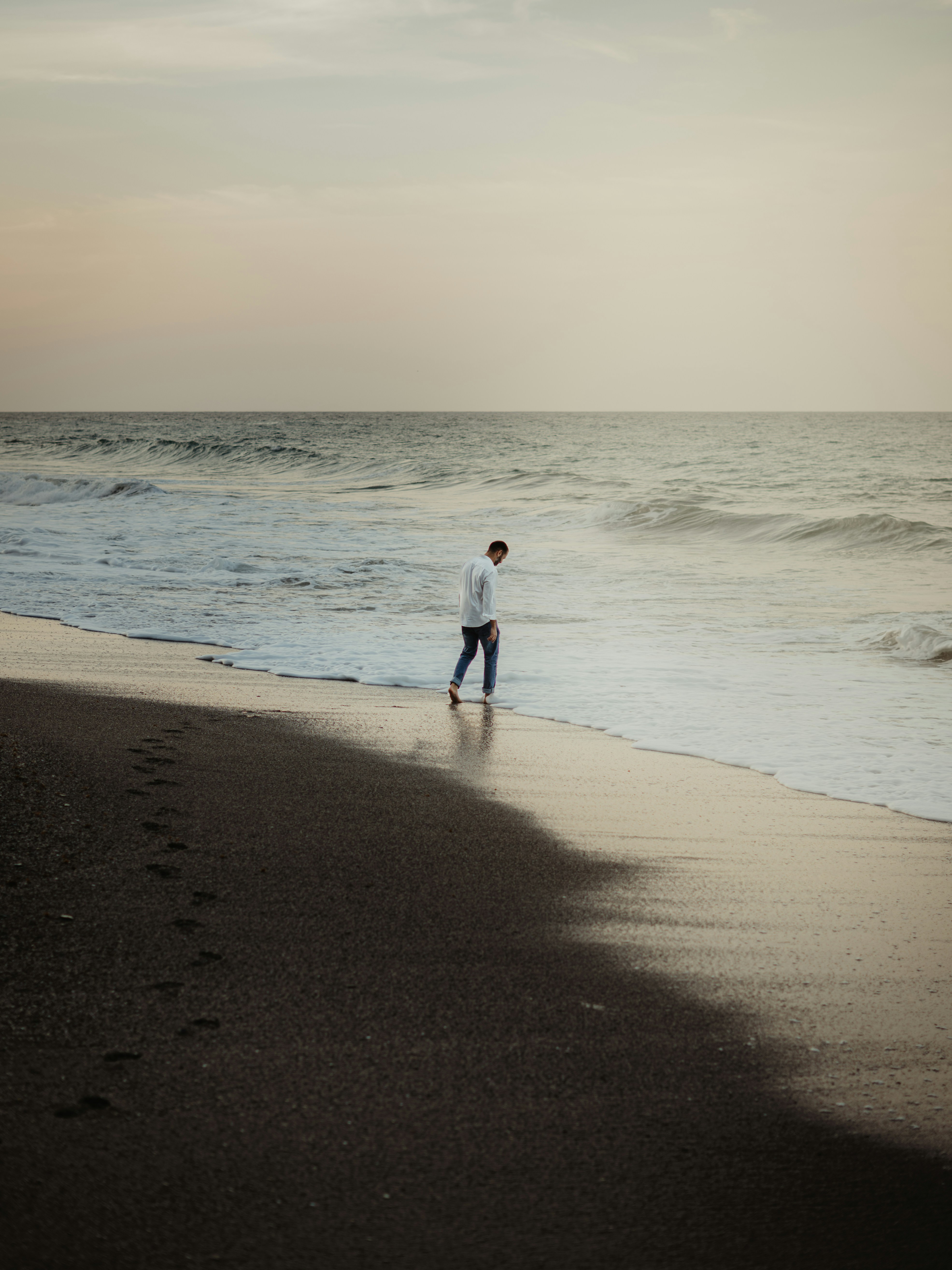 A lone figure walks along a sunlit shoreline, where gentle waves meet the dark sand, conveying calm solitude. This photographic capture focuses on the person against an expansive seascape, with a subtle horizon and soft, natural tones.