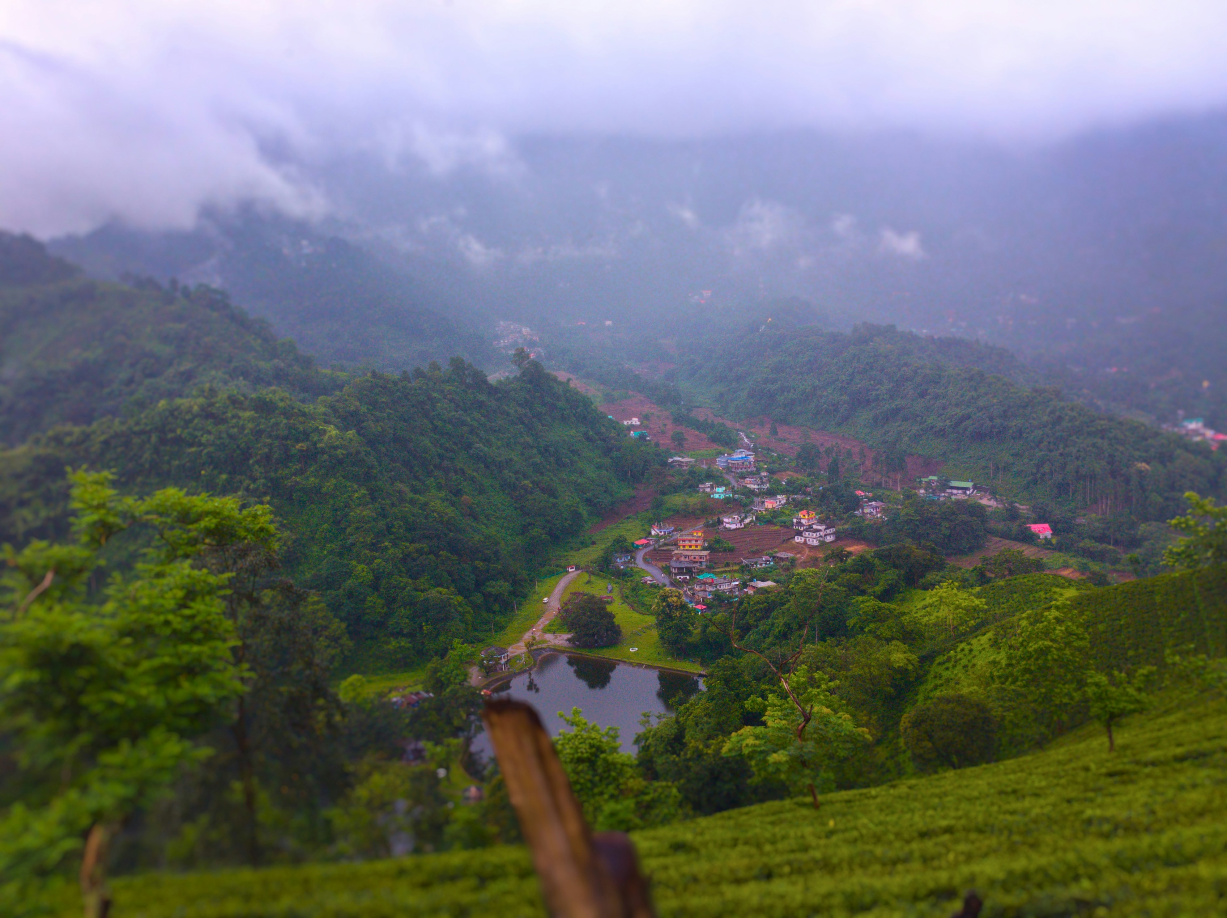 High-angle scenic view of a quiet mountain village nestled in a lush green valley, surrounded by rolling terraced tea plantations, dense forests, a serene lake, and misty clouds drifting over the hills.