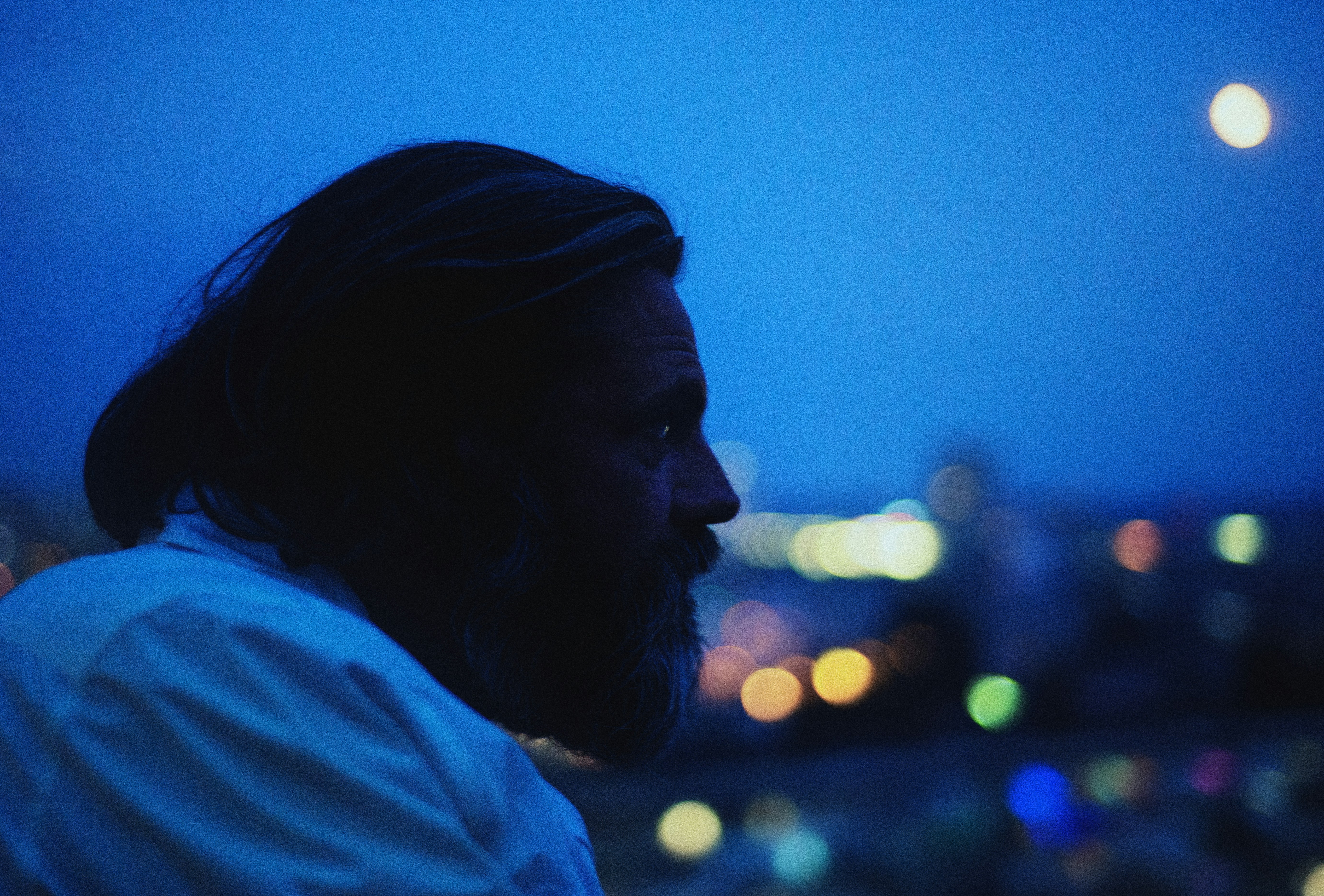 A lone figure with a beard gazes toward a distant cityscape under blue night light, the scene softly blurred with bokeh. This is a candid portrait with a moody, atmospheric backdrop.