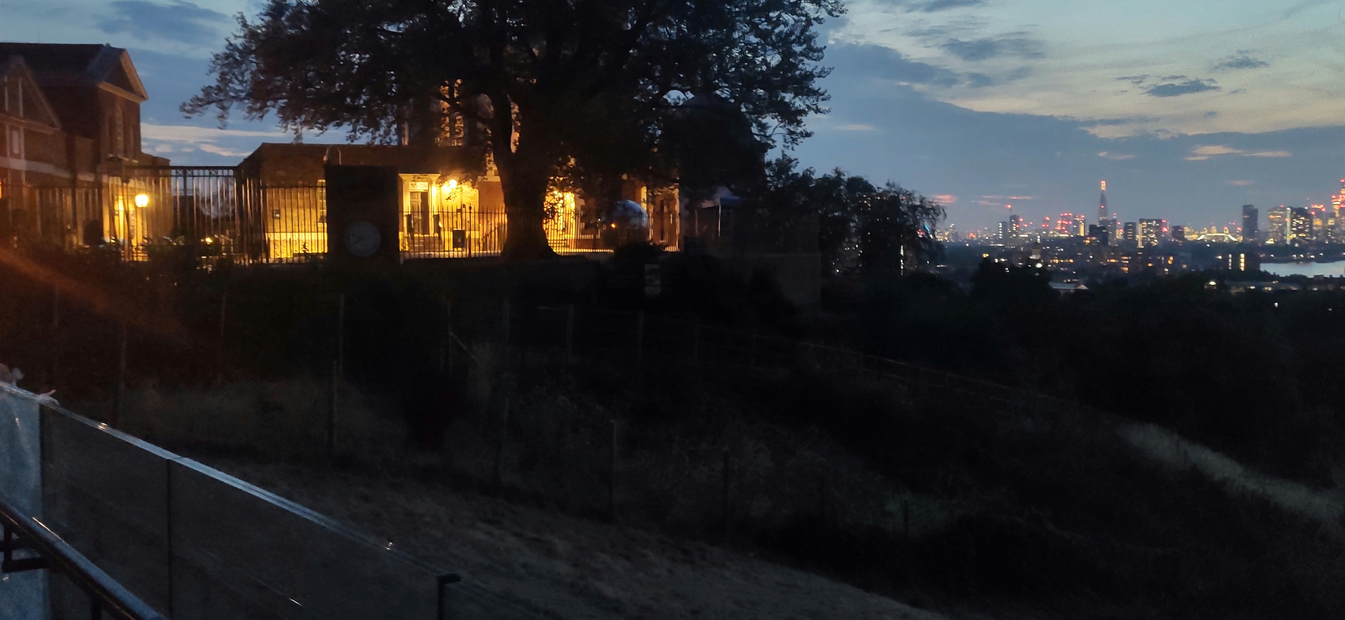 Low-light dusk scene with a lit terrace on the left and a distant city skyline across a dark hillside. The composition emphasizes the warm glow of the building against the cool urban horizon.