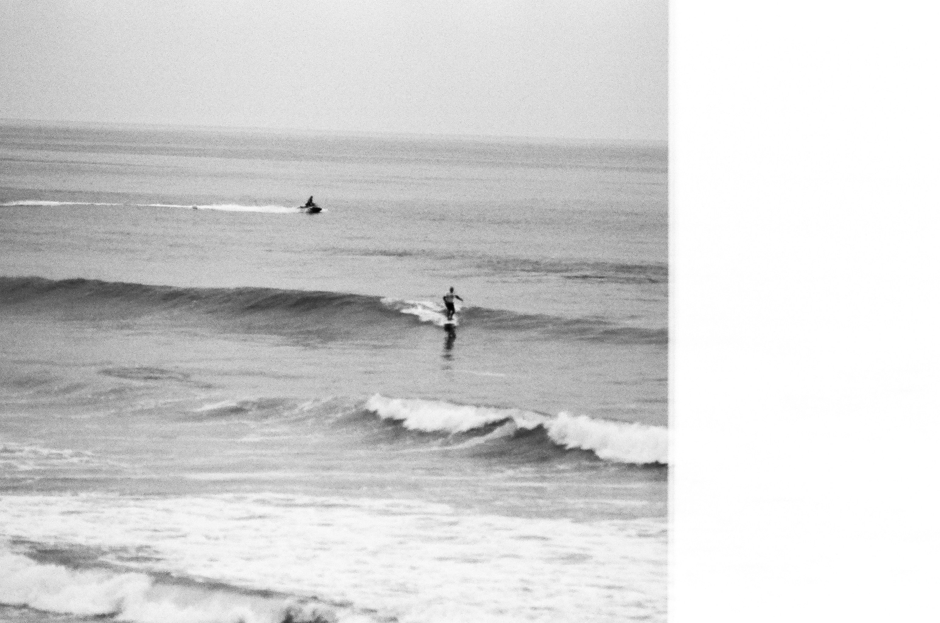 A lone surfer stands on a gentle ocean swell, captured in grayscale with soft horizon and distant watercraft in the distance.