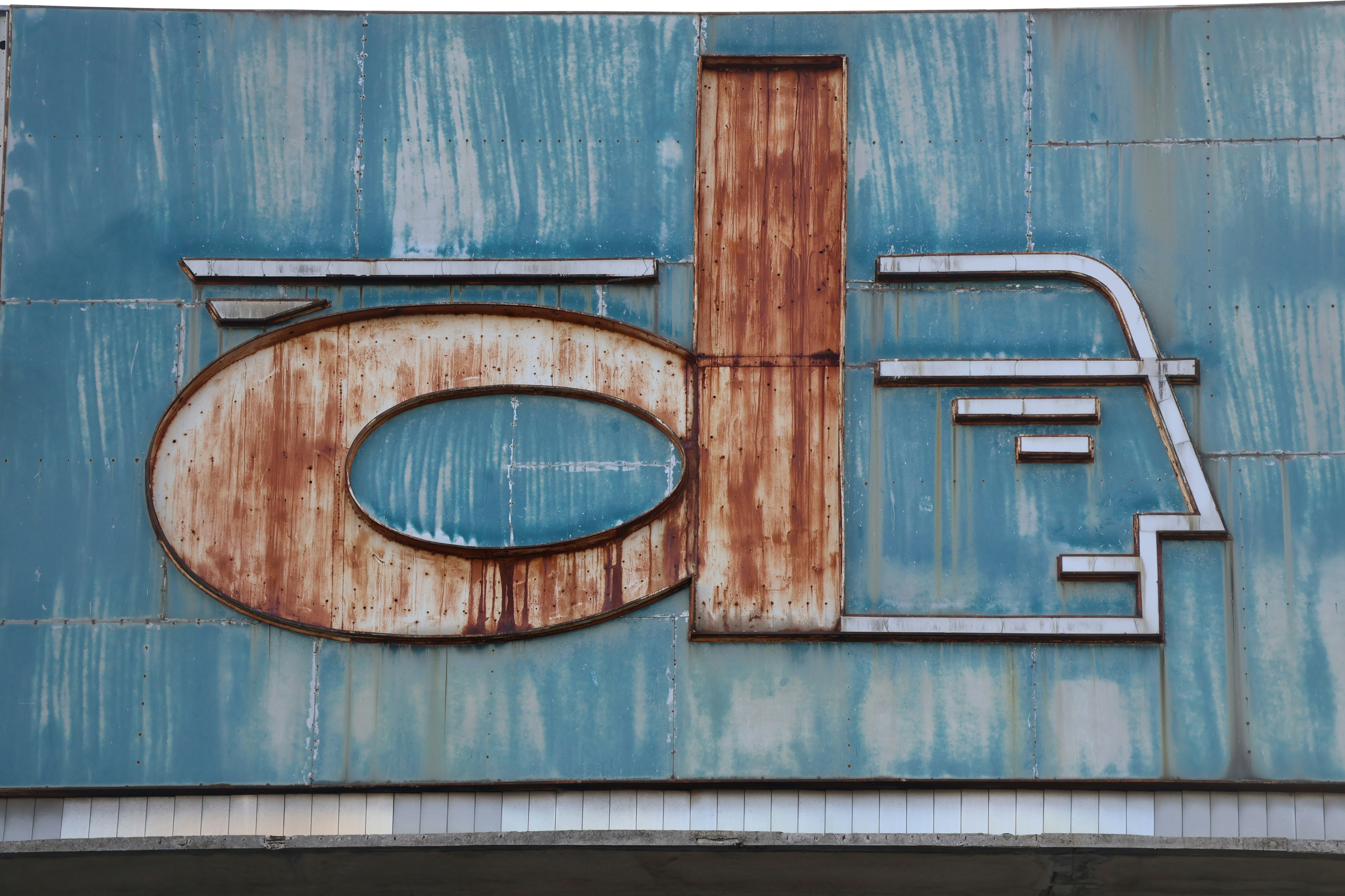 A close-up of a weathered, heavily rusted geometric metal sign mounted on a faded teal paneled wall. The peeling paint, rich rust tones, and dripping water marks create a strong industrial, moody, and vintage aesthetic