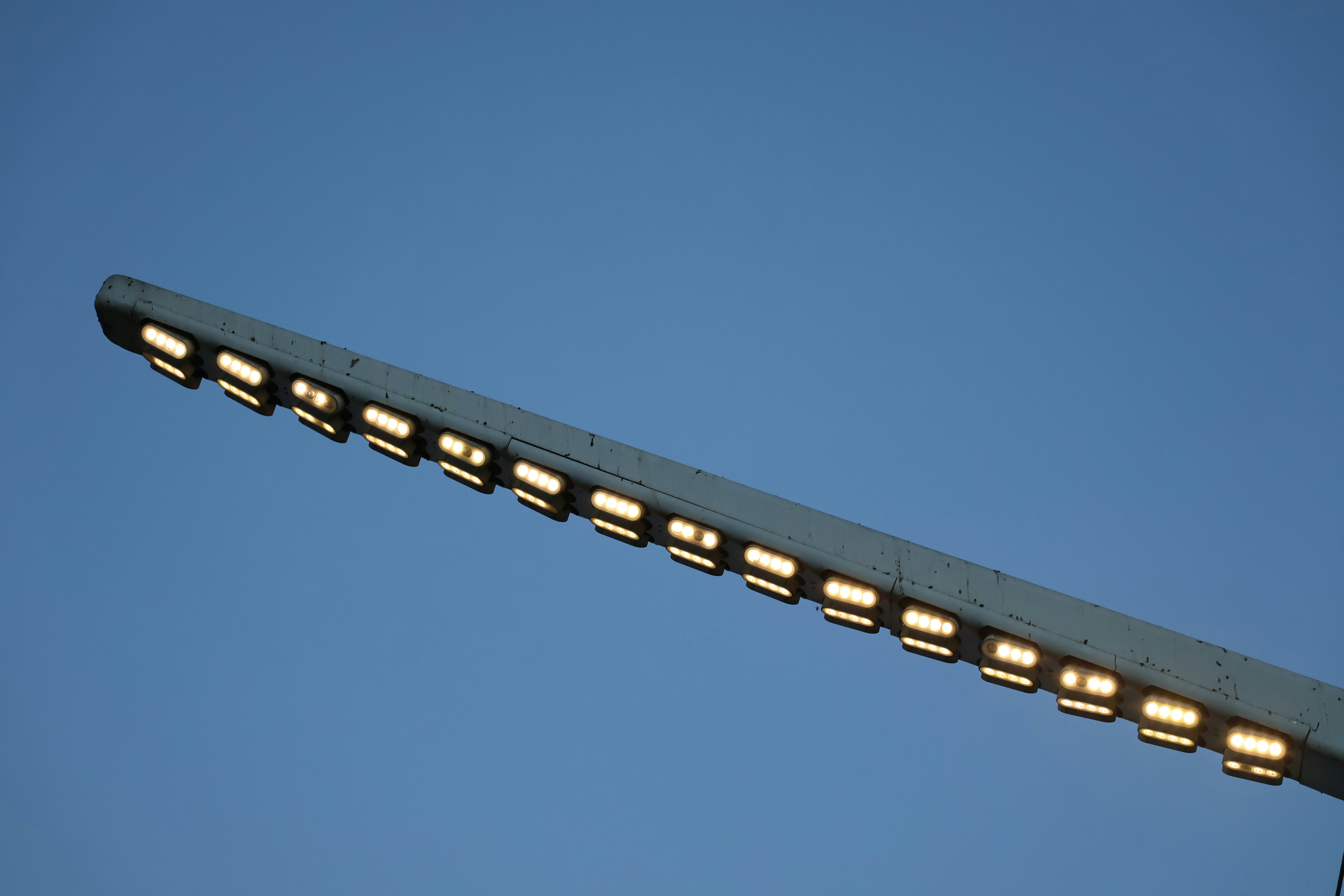 A diagonal LED streetlight strip stretches across a clear blue sky, emphasizing linear repetition and geometric contrast.
