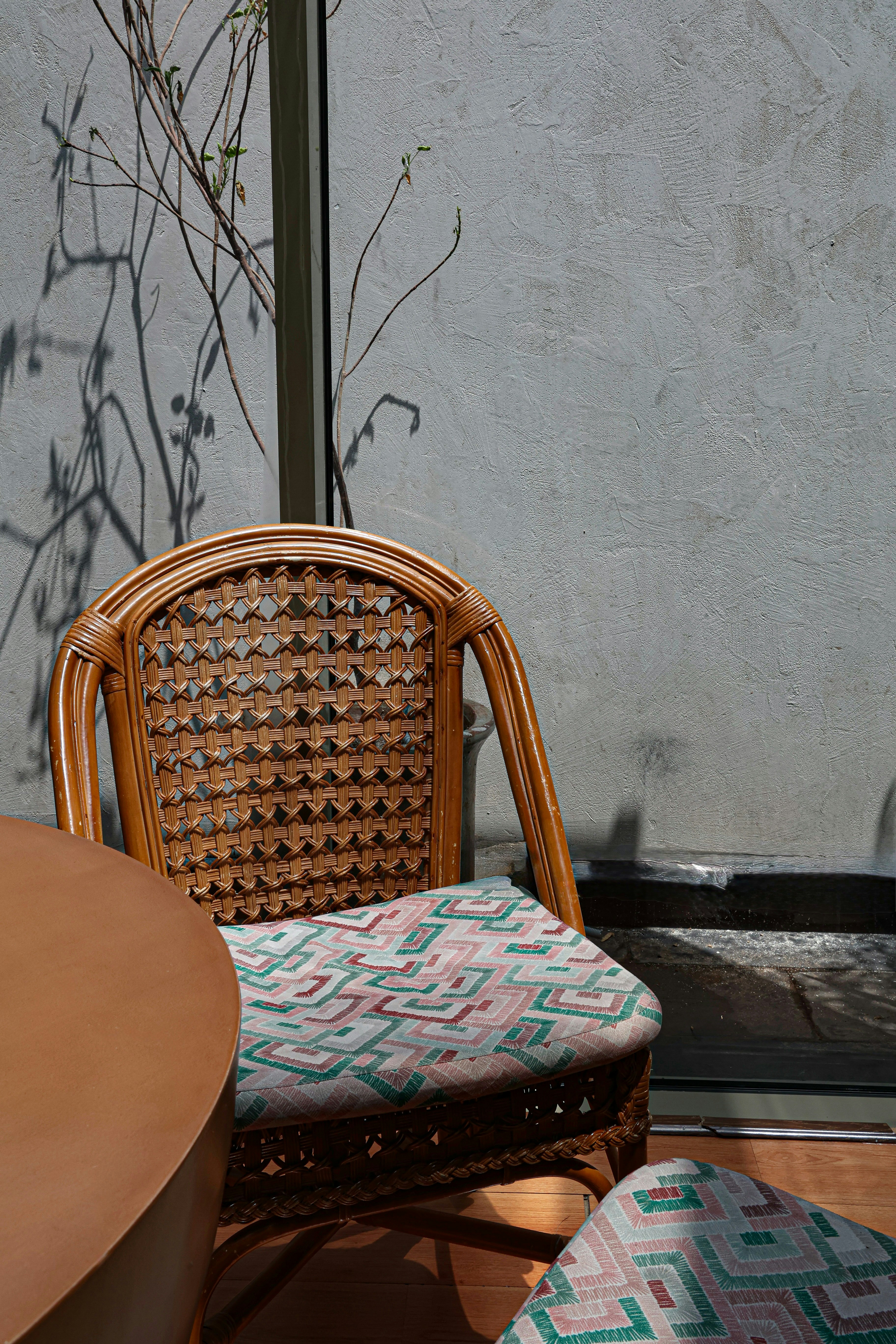 Wicker chair with a patterned cushion sits beside a round table by a sunlit window; soft shadows and a textured wall provide a calm, indoor scene. This is a staged photography of a home setting.