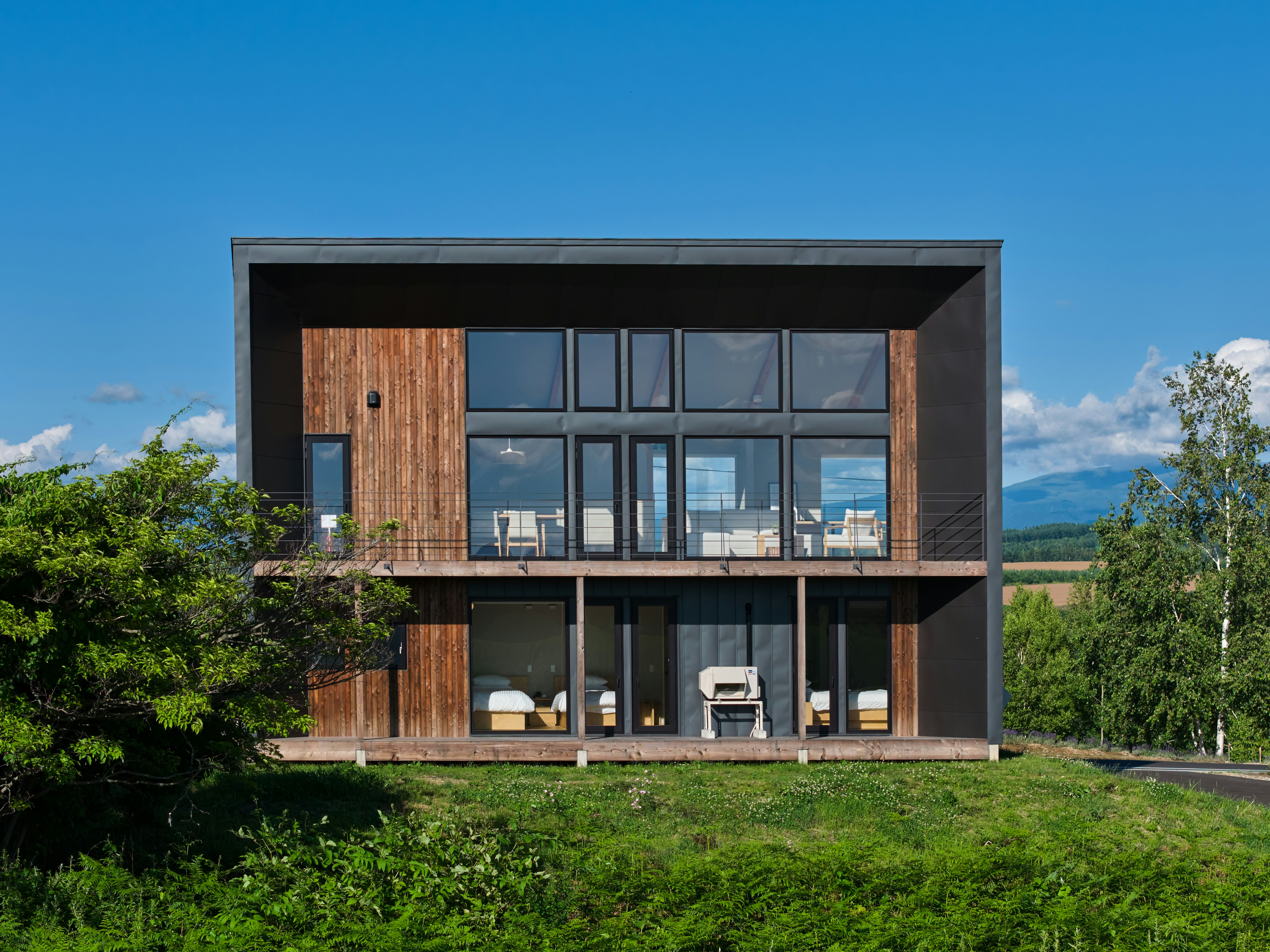 Sunlit modern house with a timber façade sits on green lawn under a clear blue sky; large windows reveal interior seating and a bright, open living space.