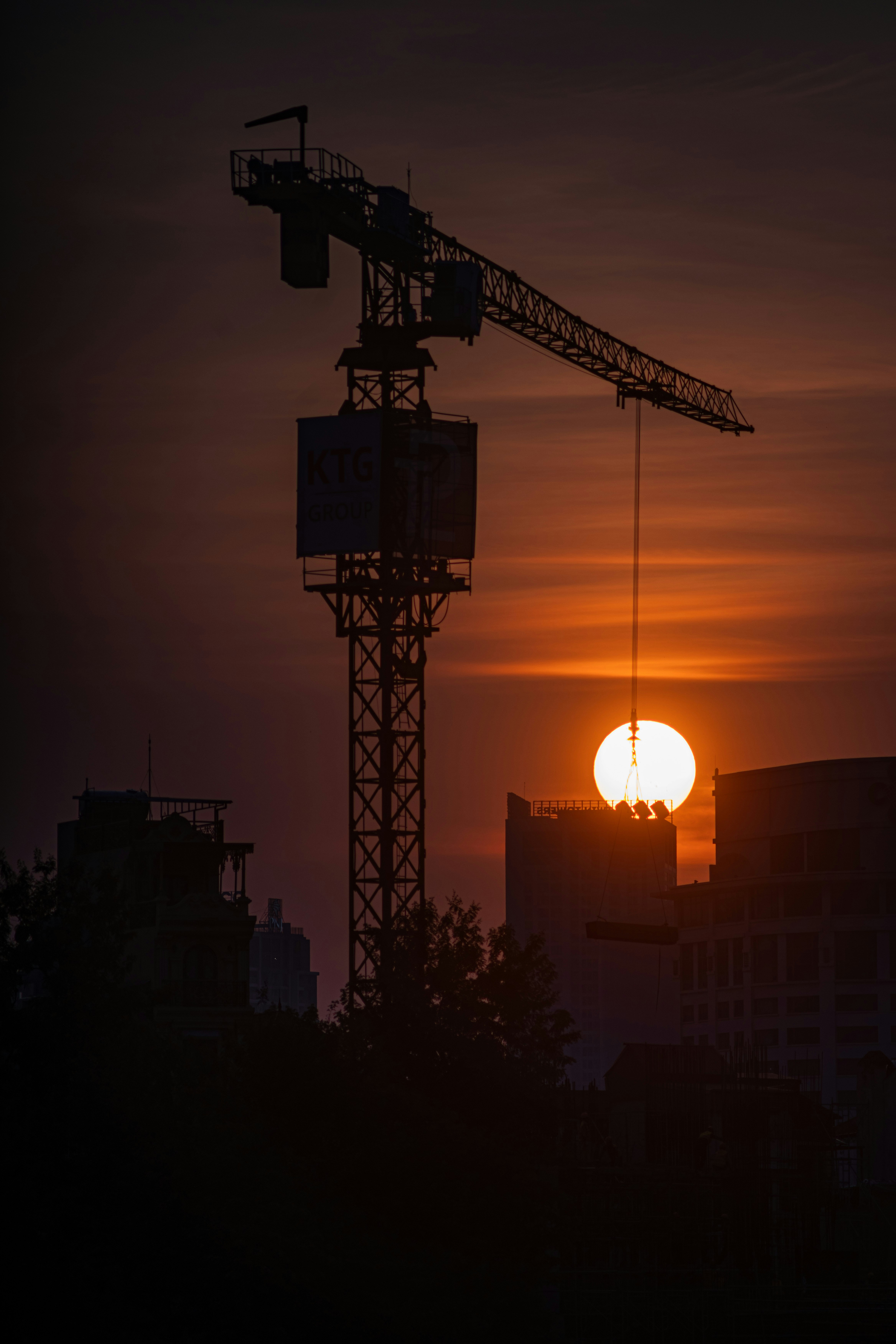 Crane silhouette against a fiery sunset over a city skyline, with the sun poised near the crane’s hook and buildings in shadow. Photograph captures industrial scale and warm horizon tones.