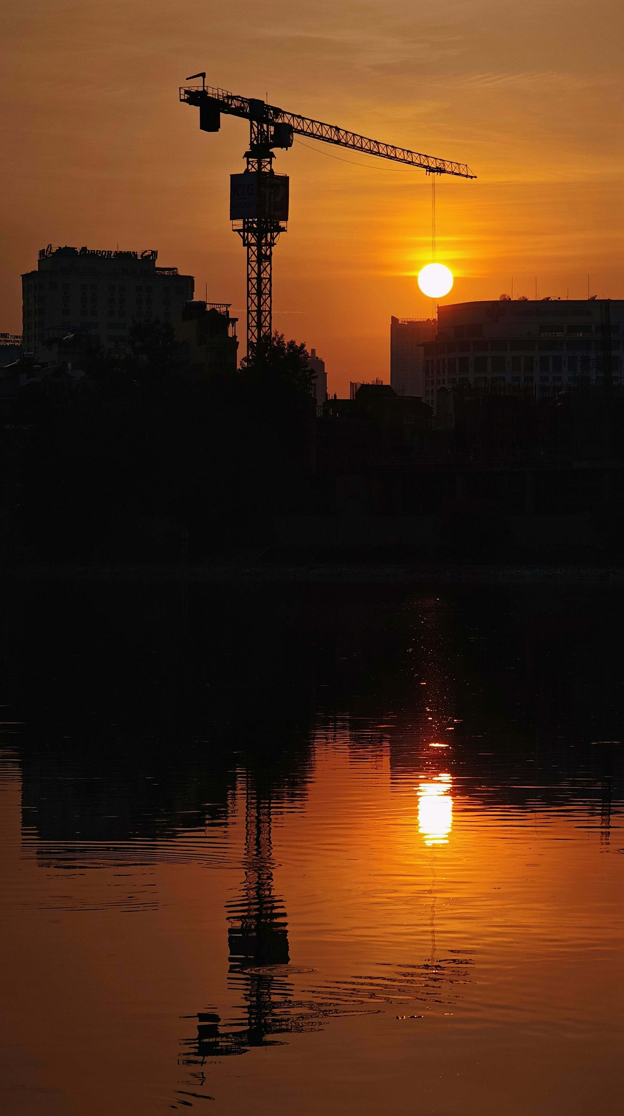 Crane and skyline at sunset above a dark cityscape, with warm orange reflections on calm water. The image captures the silhouette and the sun aligned with the crane, highlighting urban activity and reflective surface textures.