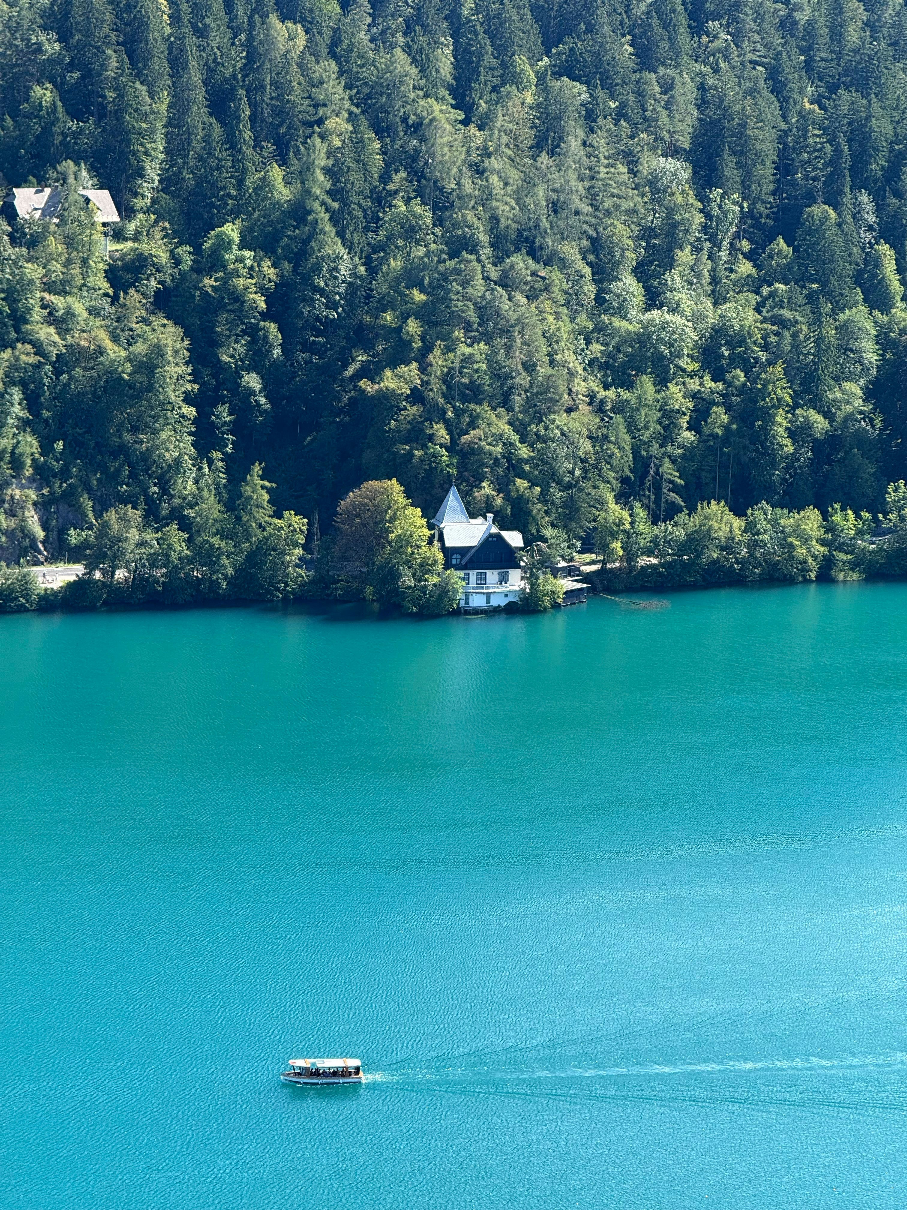 A turquoise lake fringed by dense evergreen forest with a small lakeside house and a boat gliding across the water.