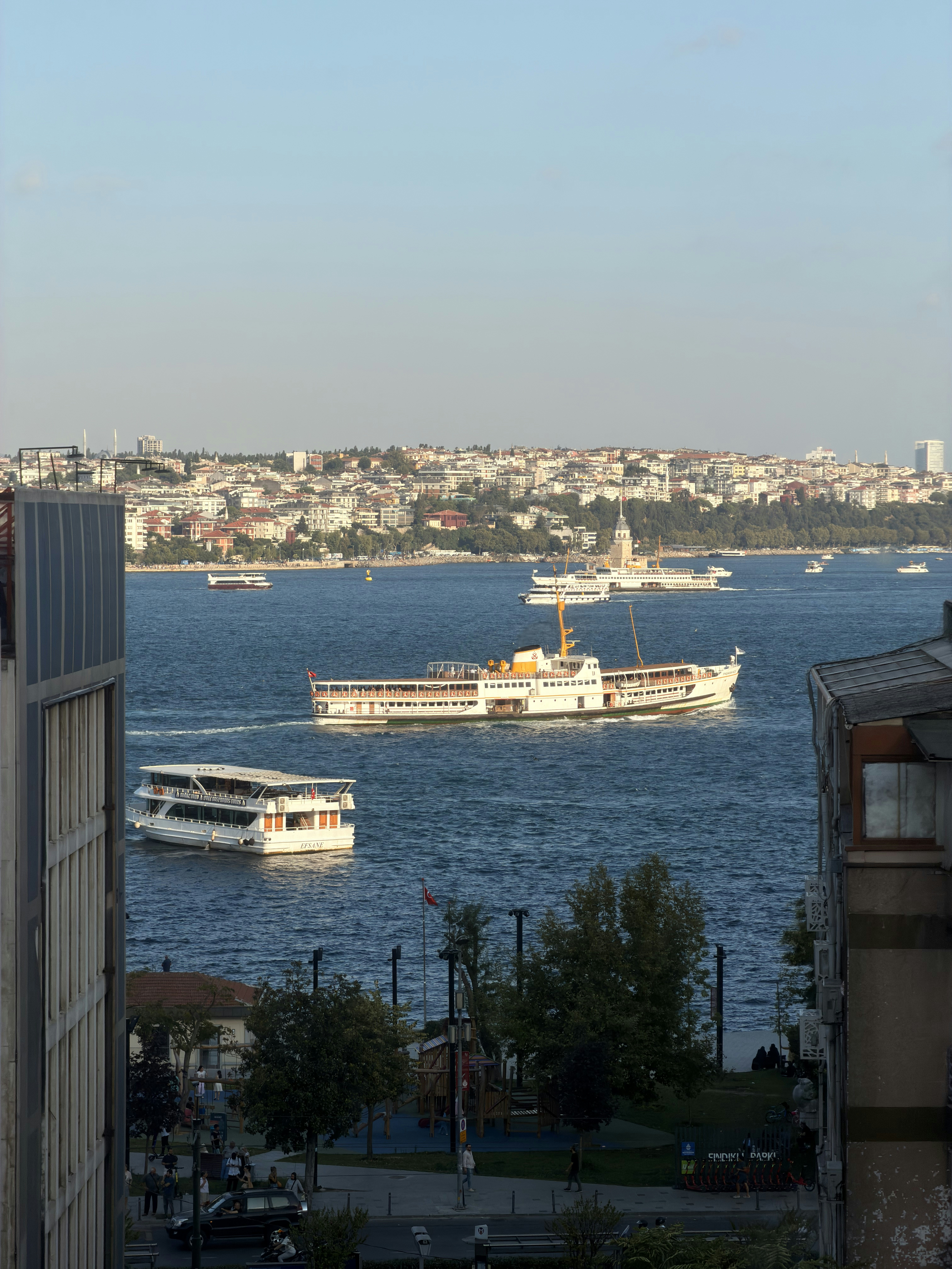 A harbor view with ferries gliding across a blue strait, framed by buildings on either side and a park in the foreground.