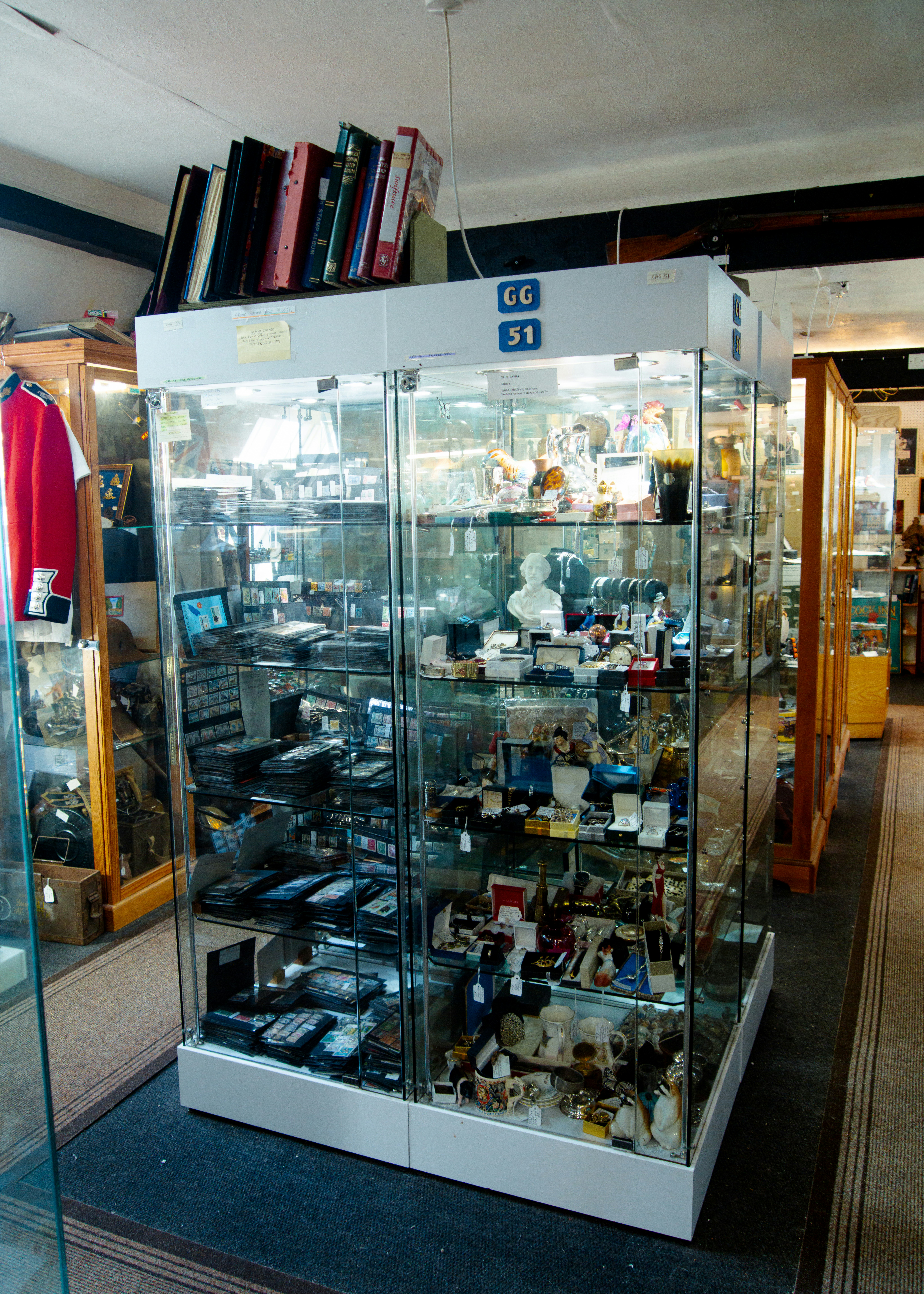 A glass display cabinet filled with assorted vintage electronics and trinkets sits in a cluttered shop, with books stacked on top and warm ambient lighting highlighting collectibles inside.