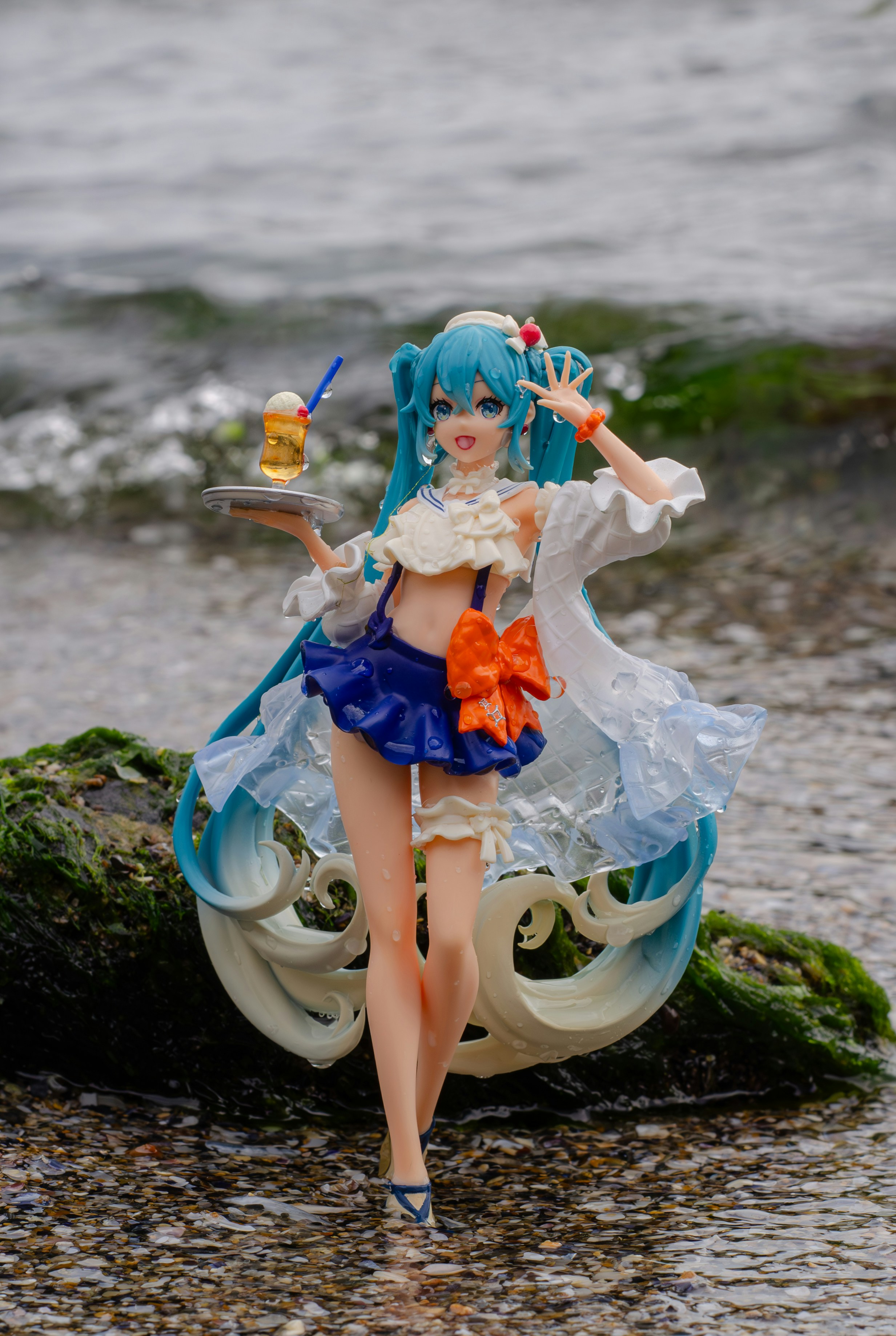 Colorful anime figure posed at the water's edge with a drink on a tray, set against rocky, mossy shoreline.