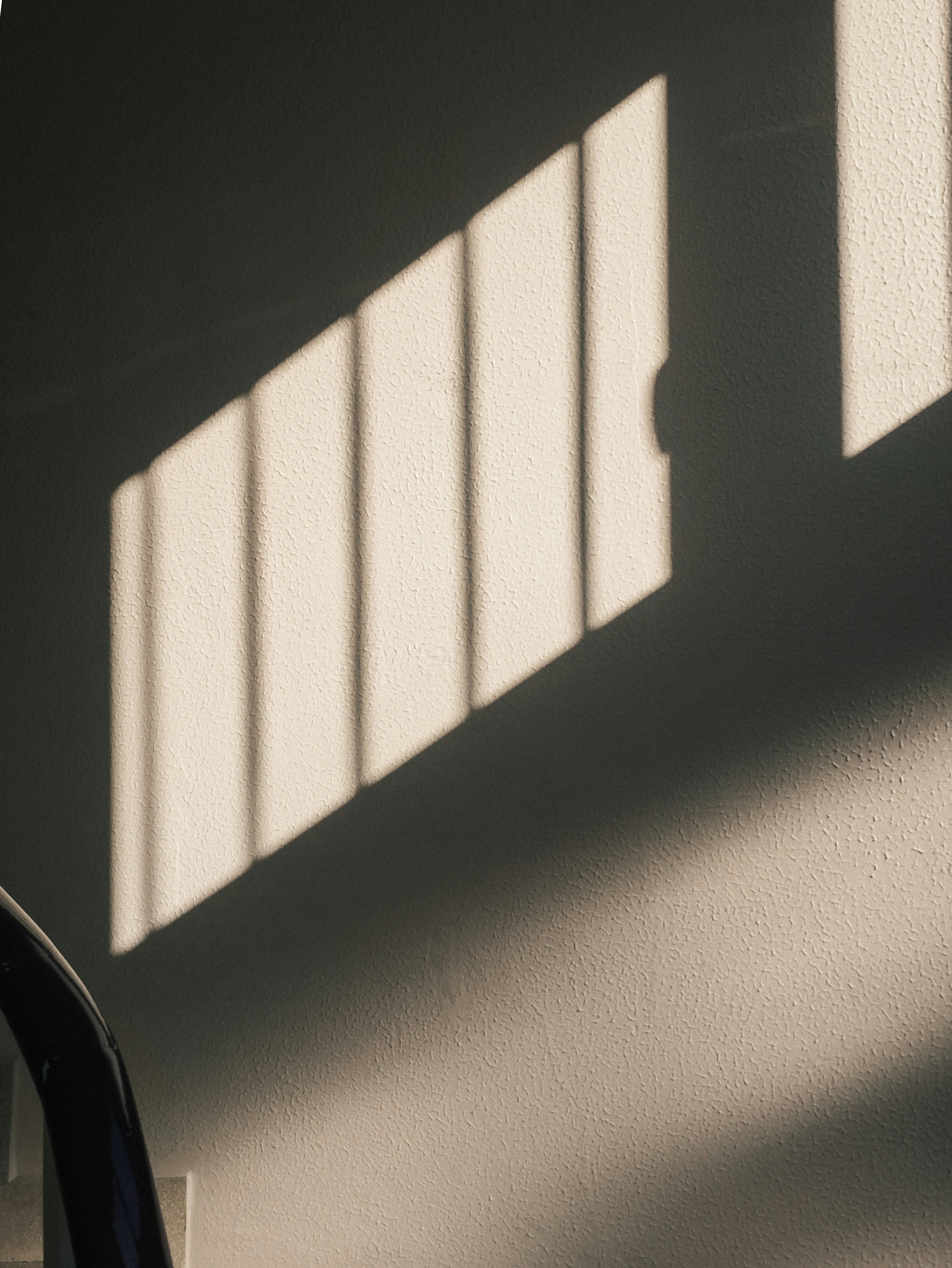Diagonal window shadow casts bold rectangular bars across a textured wall, with a hint of a chair edge in the lower-left corner.