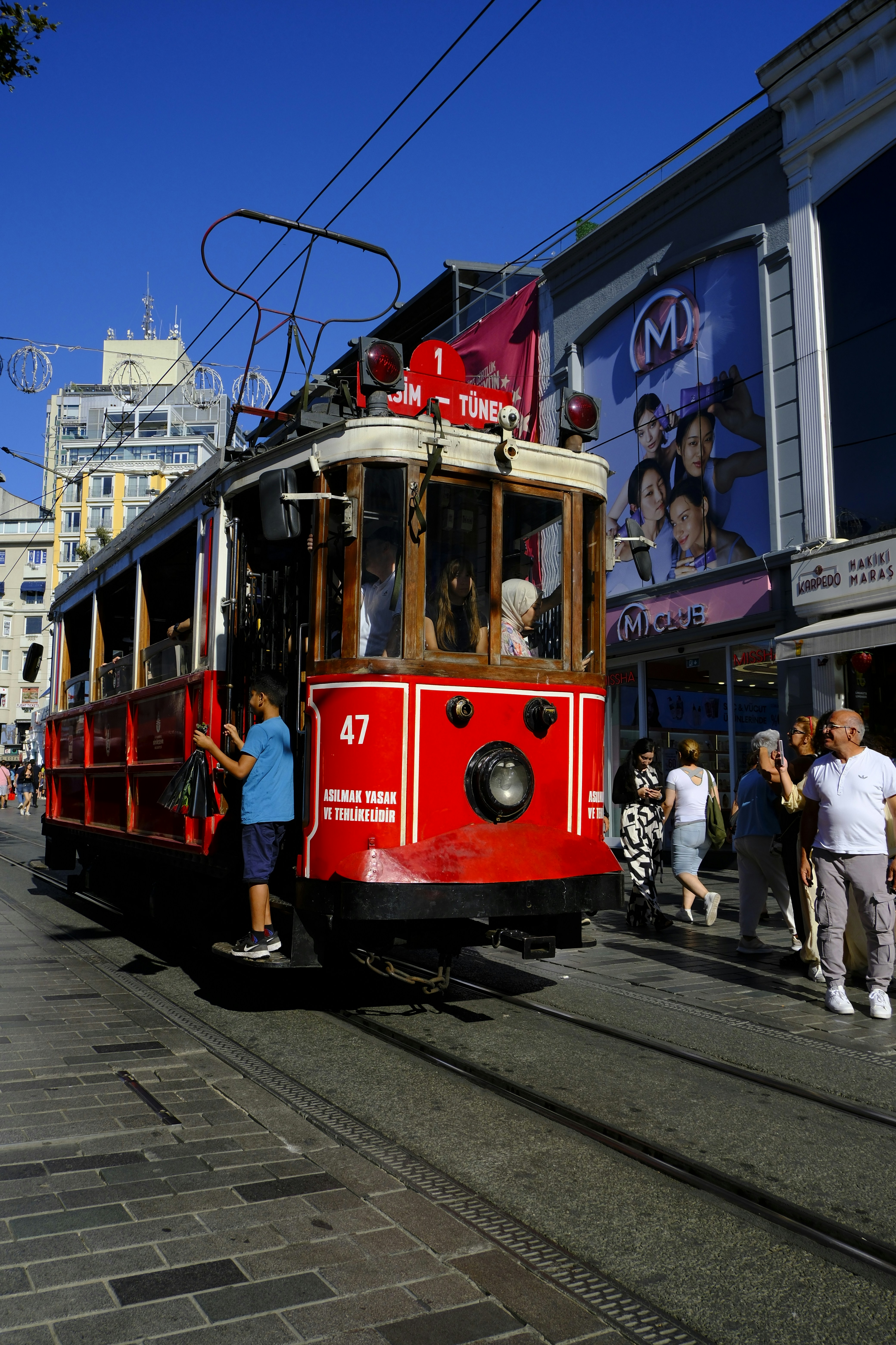 A vintage red tram anchors a bustling urban scene with pedestrians along a storefront-lined street under a bright blue sky.