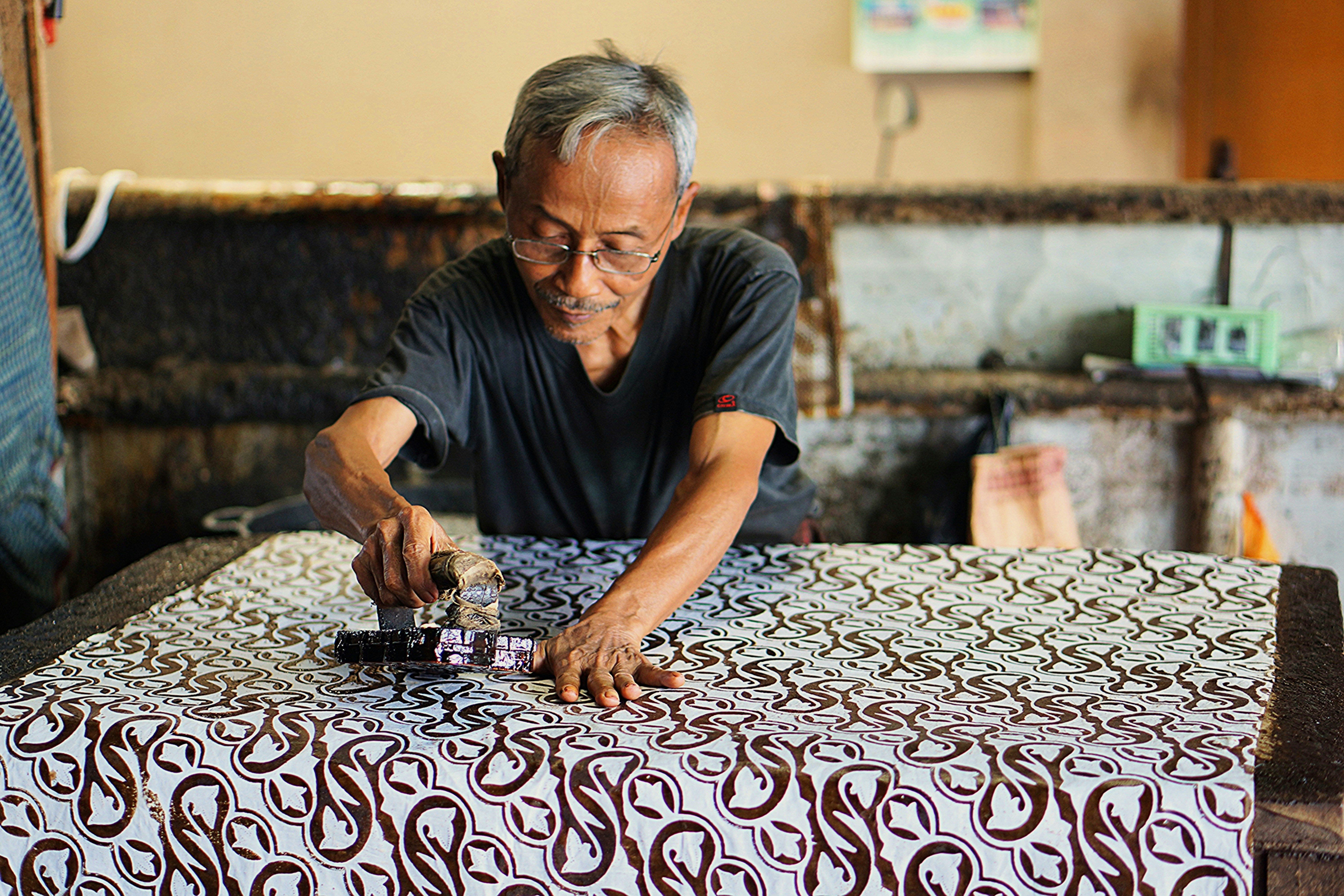 patterned fabric laid out on a worktable as an elderly craftsman carves a tool, focused on his meticulous hands and the textile design.