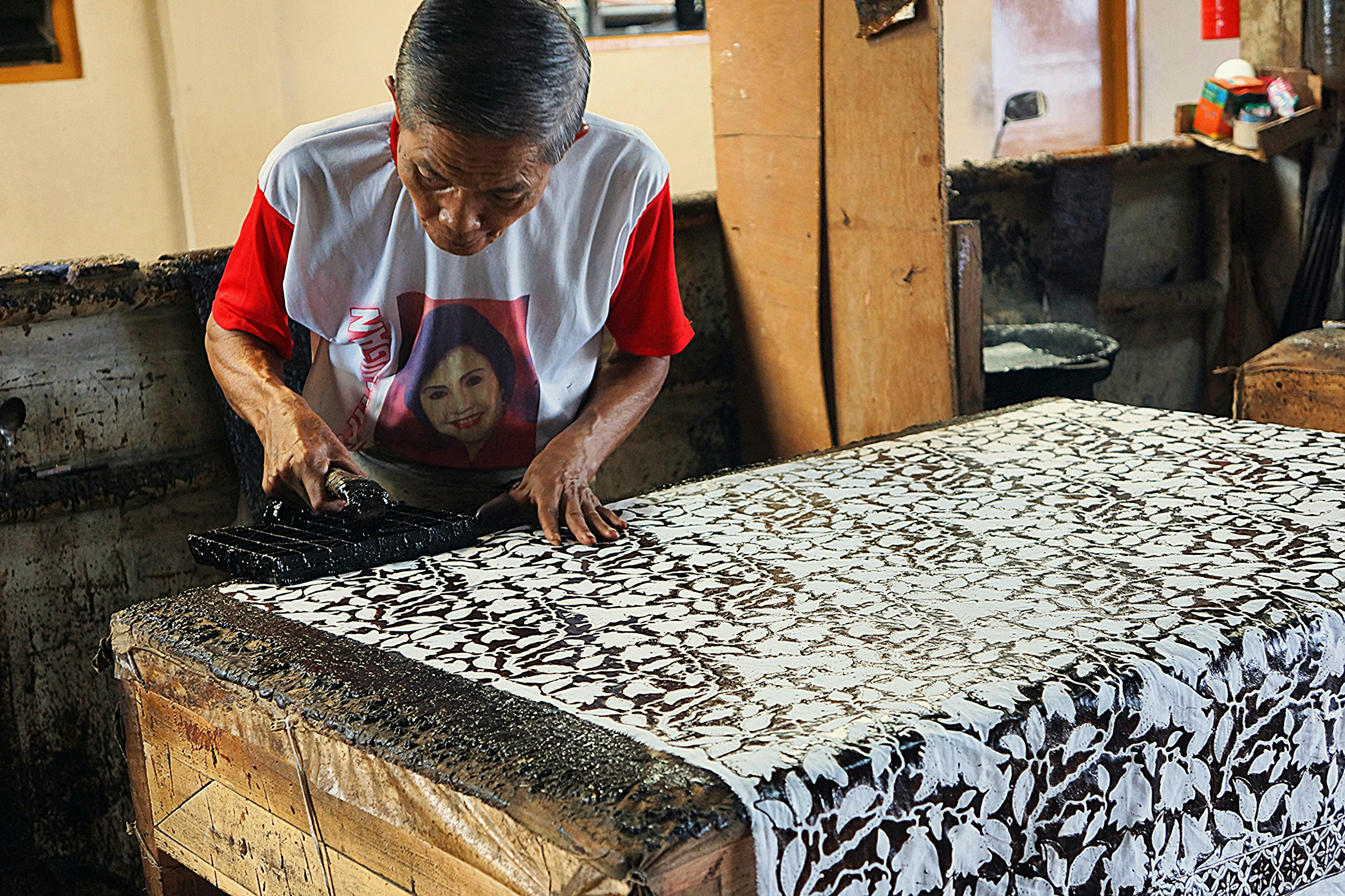 Traditional Batik Printing by Hand Description: A documentary photograph showing a skilled artisan carefully applying dye to a large piece of batik fabric using a traditional printing tool. The intricate black-and-white floral pattern spreads across the cloth, highlighting the precision, patience, and craftsmanship involved in Indonesian batik production.