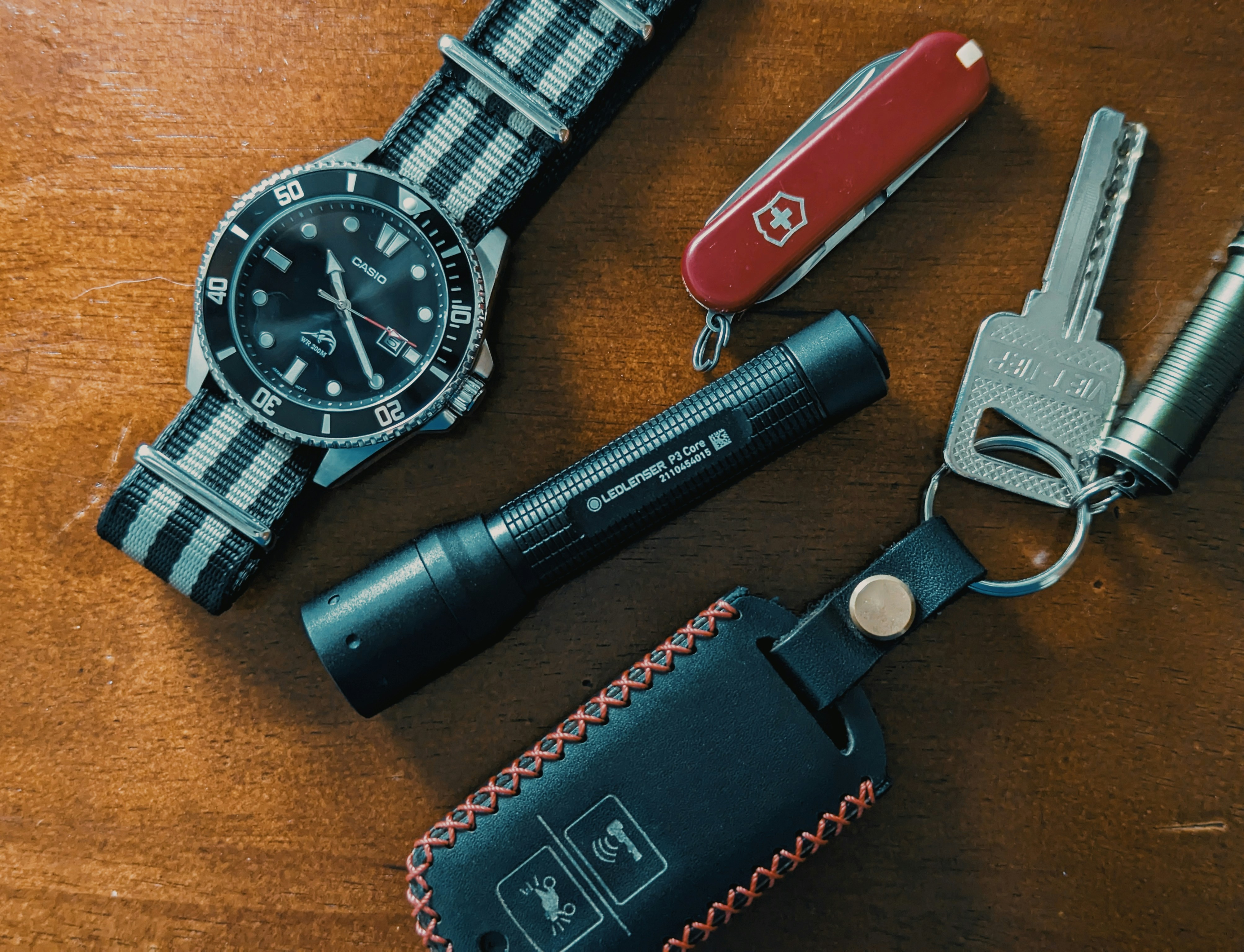 A collection of everyday carry items laid on a wooden surface, featuring a black dive watch, a compact flashlight, a red Swiss Army knife, and a leather-key fob with two keys. The arrangement highlights metallic textures and utility gear in a warm-toned setting.