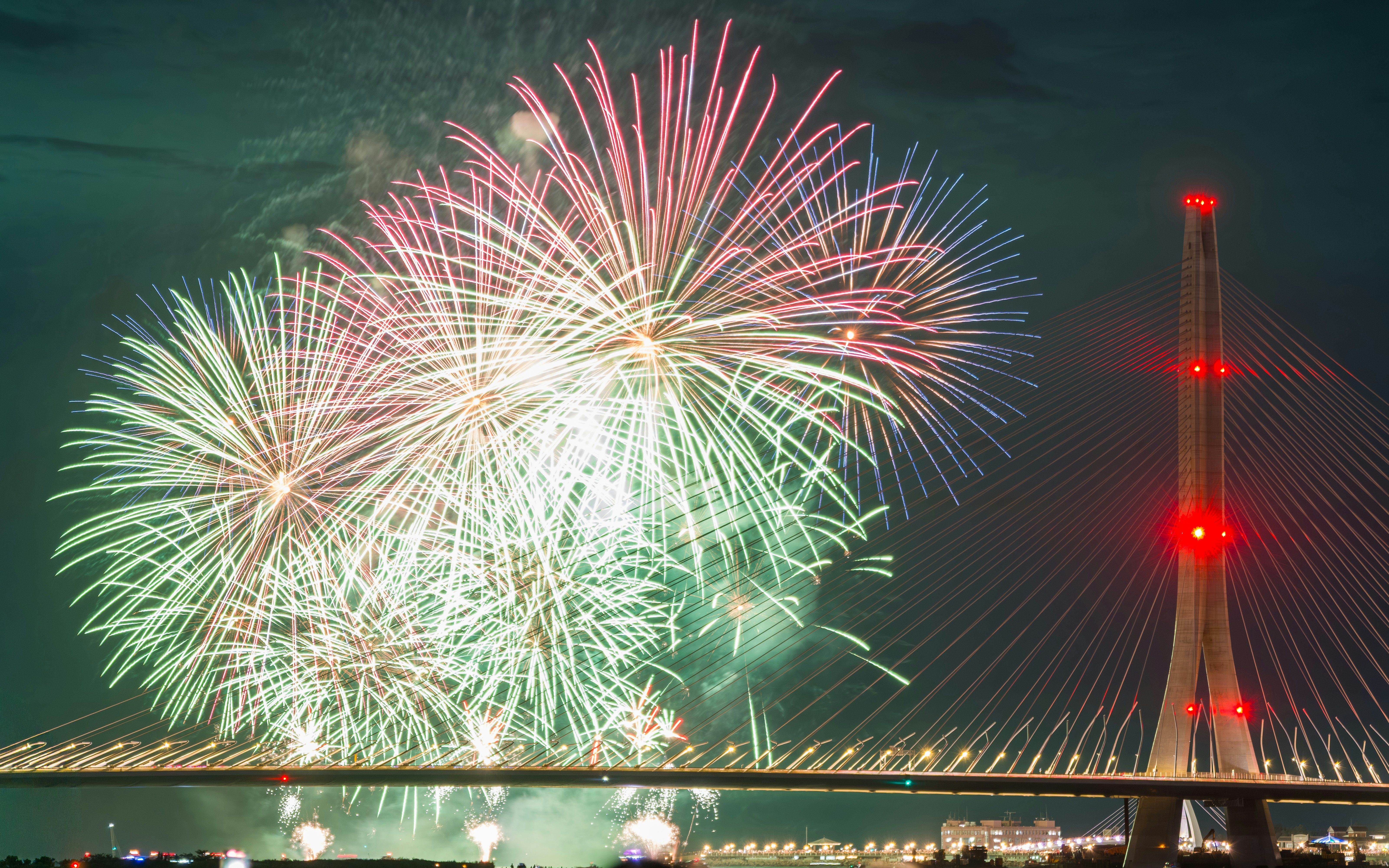 Green and red fireworks exploding above a cable-stayed bridge at night