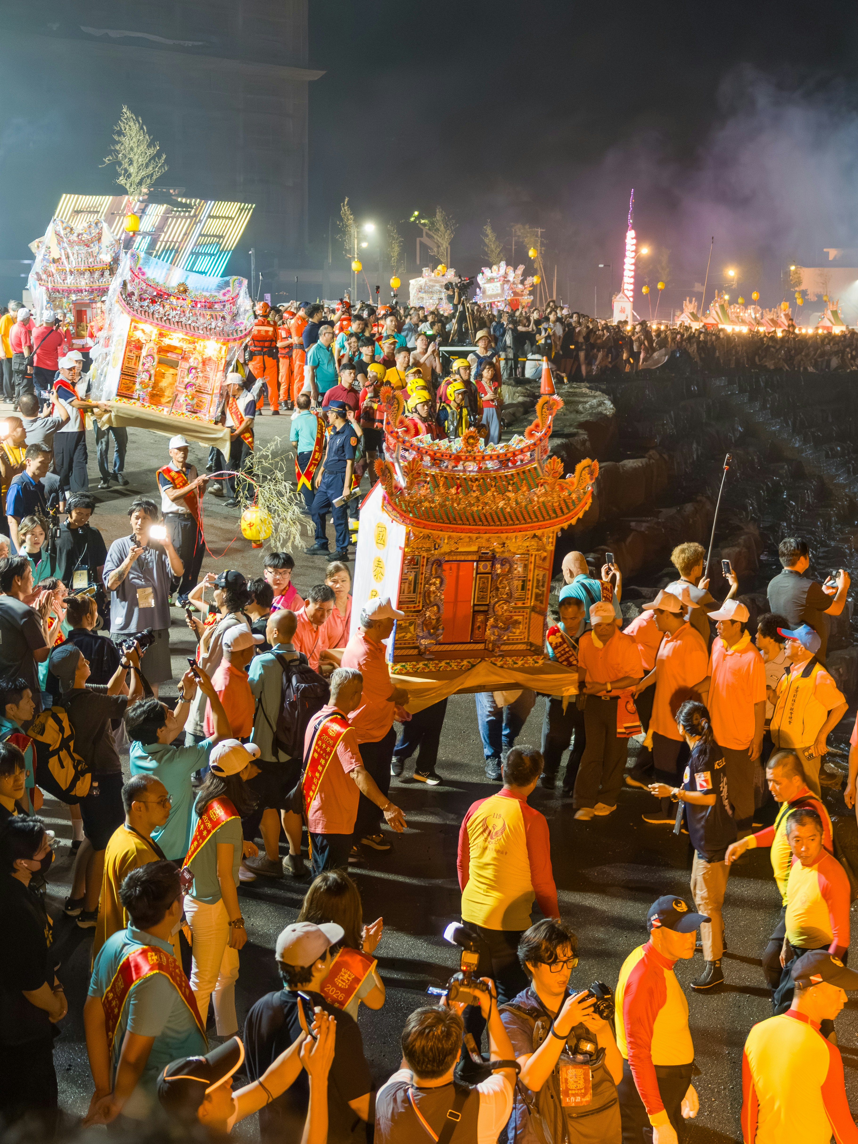 Colorful procession with ornate temple floats surrounded by musicians and spectators at a nighttime festival.