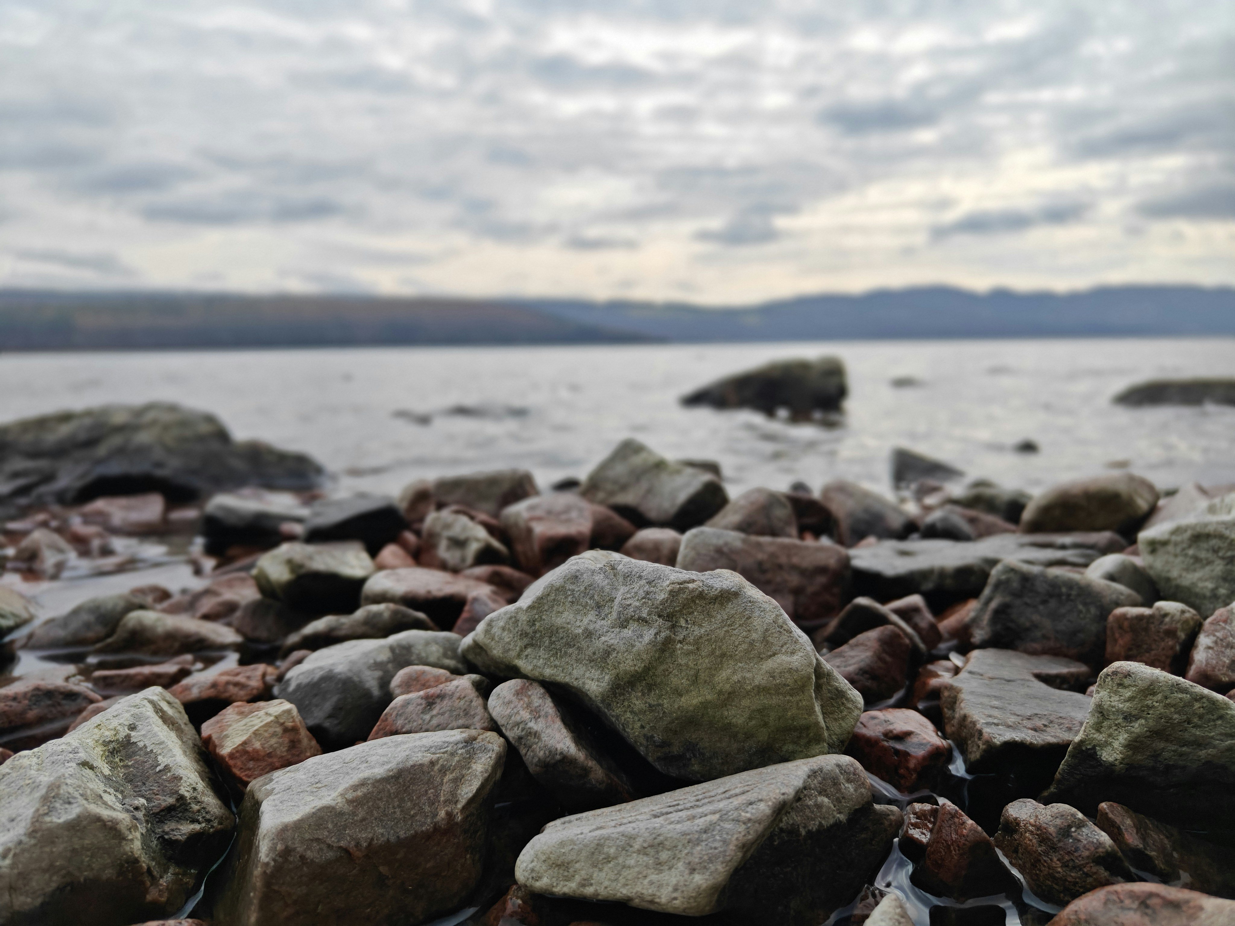 Strewn rocks along a calm lakeshore with a distant horizon and cloudy sky, highlighting texture and color of the pebbles. The scene is a natural landscape, with no people or logos visible.