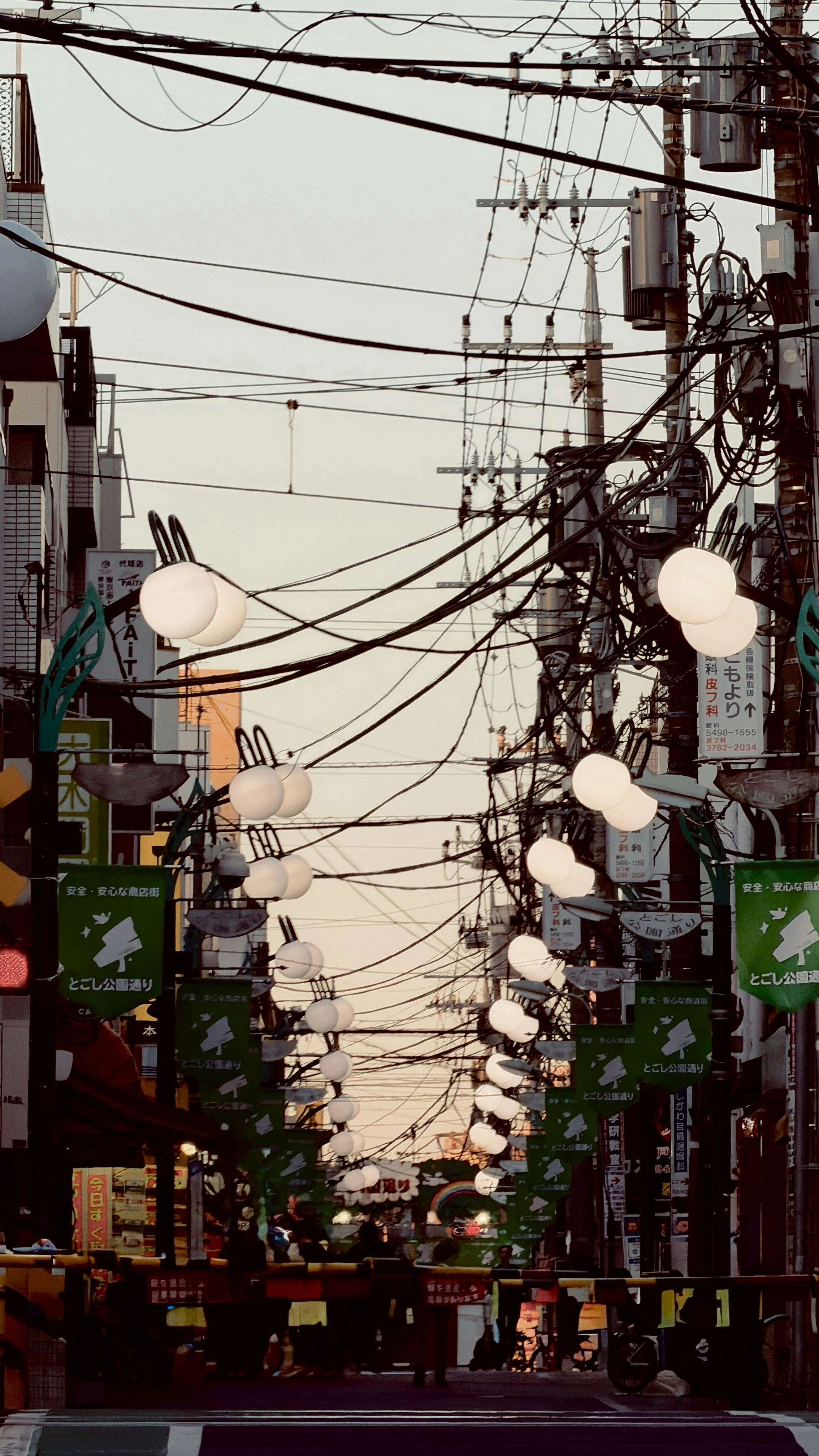 A narrow urban street drenched in warm evening light, dotted with hanging lanterns and tangled utility lines, guiding the eye toward a distant vanishing point. This is a street scene photograph with real-world signage and storefronts.
