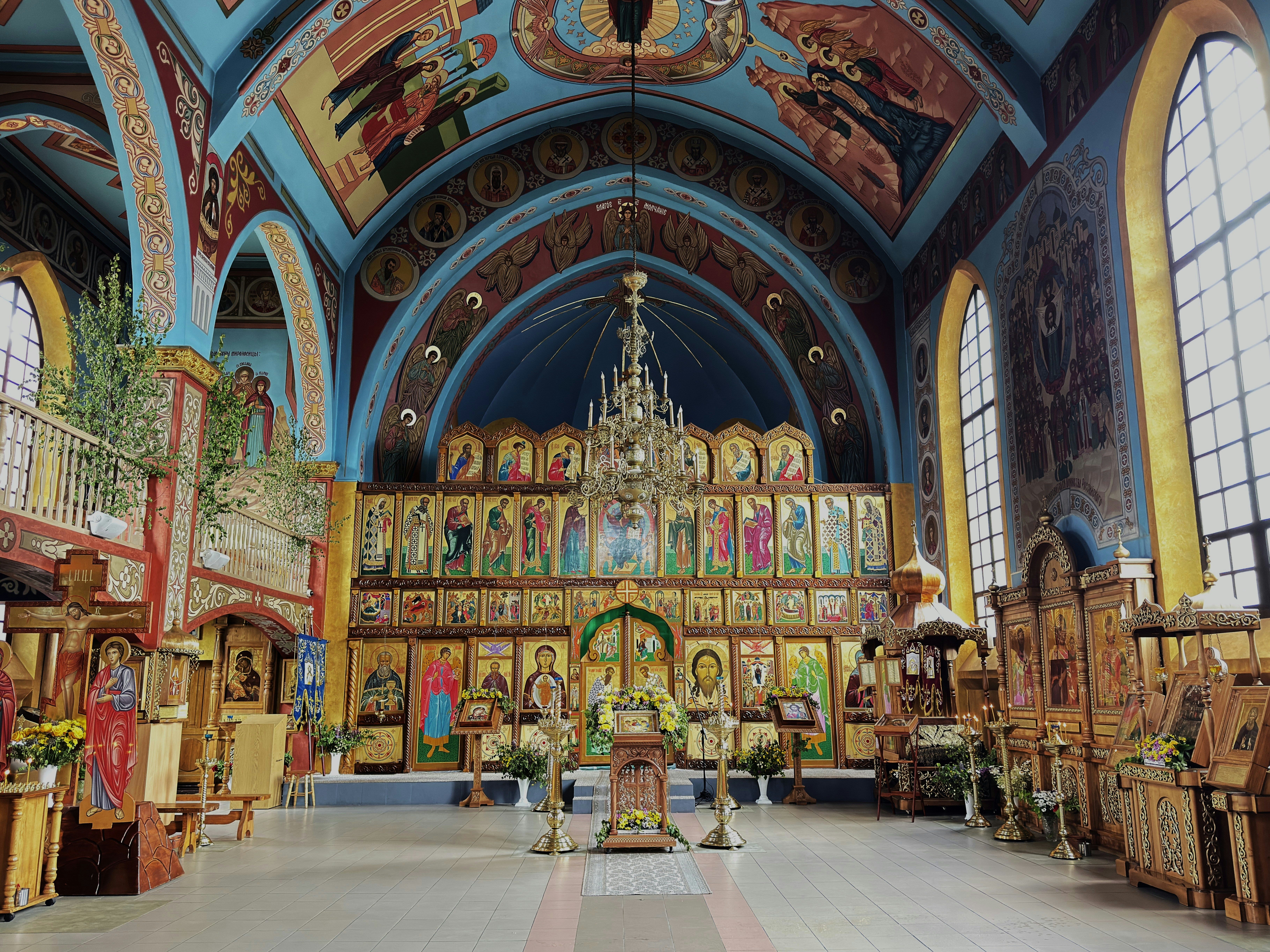 Detailed iconostasis inside a church with multiple gilded icons and ornate woodwork, centered on an altar framed by a tiered icon screen and a tall, intricate chandelier.