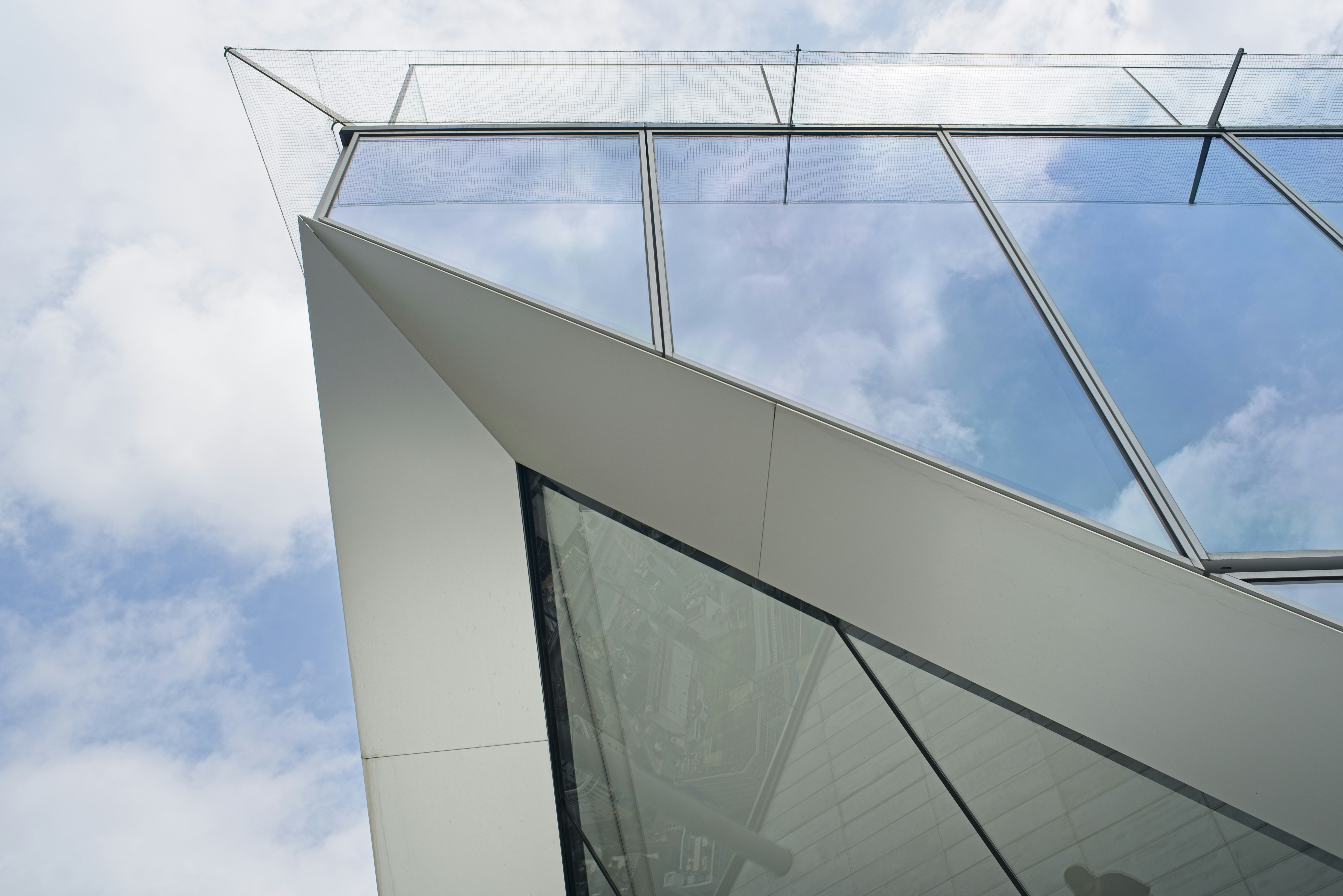 A striking low-angle view showcases the bold, angular white architecture of Shibuya Sky against a partly cloudy sky, netted safety barriers stretching along the rooftop's edge. Reflected in the sloped glass panel below, a faint aerial cityscape appears distorted, adding an unexpected layer of depth to the sharp geometric composition. The clean lines and minimalist palette capture the striking modern design of Tokyo's rooftop observation deck from a dramatic architectural perspective on a hot July afternoon.