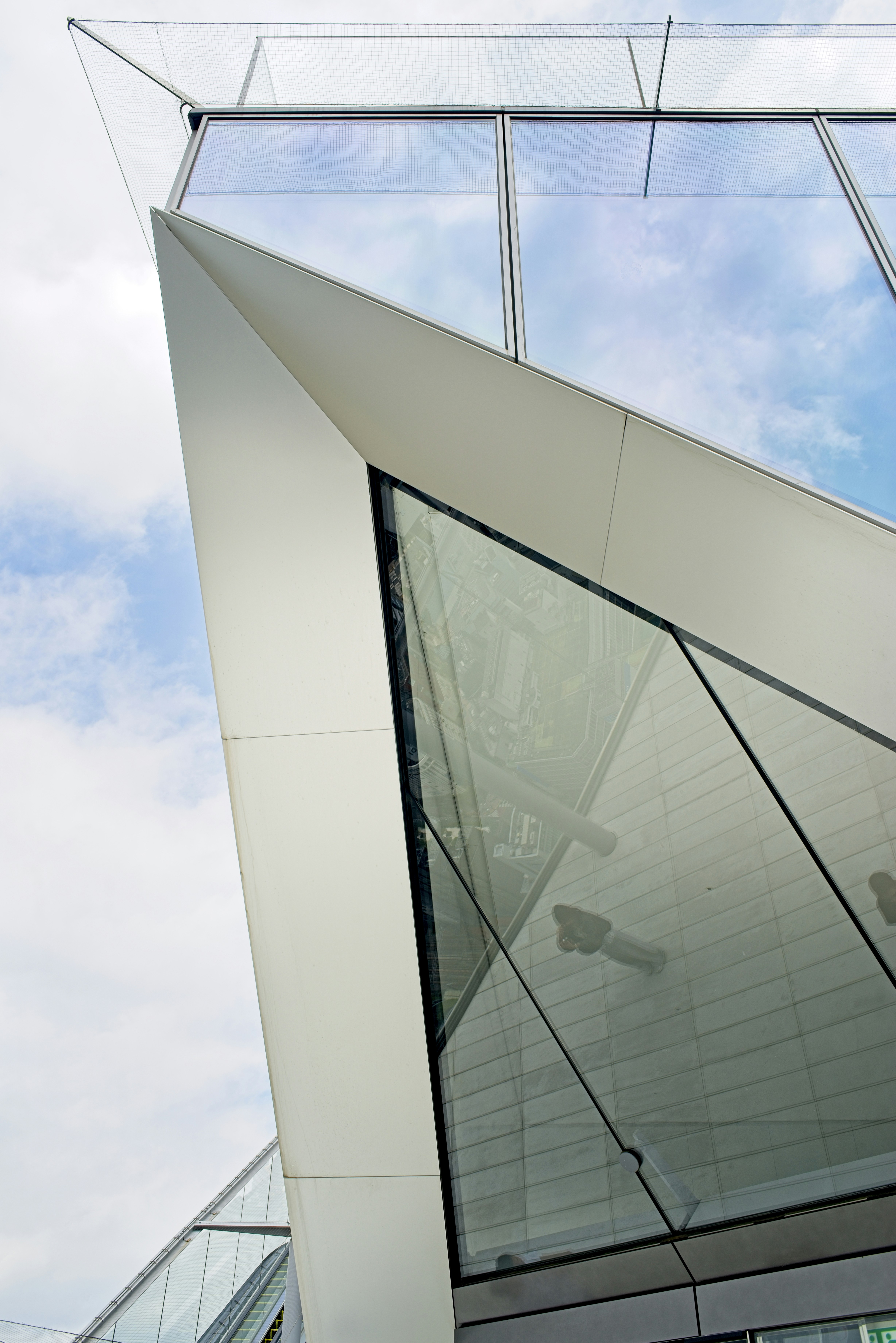 A dramatic low-angle view captures the sharp, sculptural white panels of Shibuya Sky's rooftop architecture cutting against a partly cloudy blue sky, with netted safety barriers stretching overhead. Reflected in the angled glass below, a distant aerial cityscape appears faintly visible, adding depth and complexity to the composition. The interplay of bold geometric lines, reflective surfaces, and open sky captures the striking modern design of Tokyo's rooftop observation deck on a hot July afternoon.