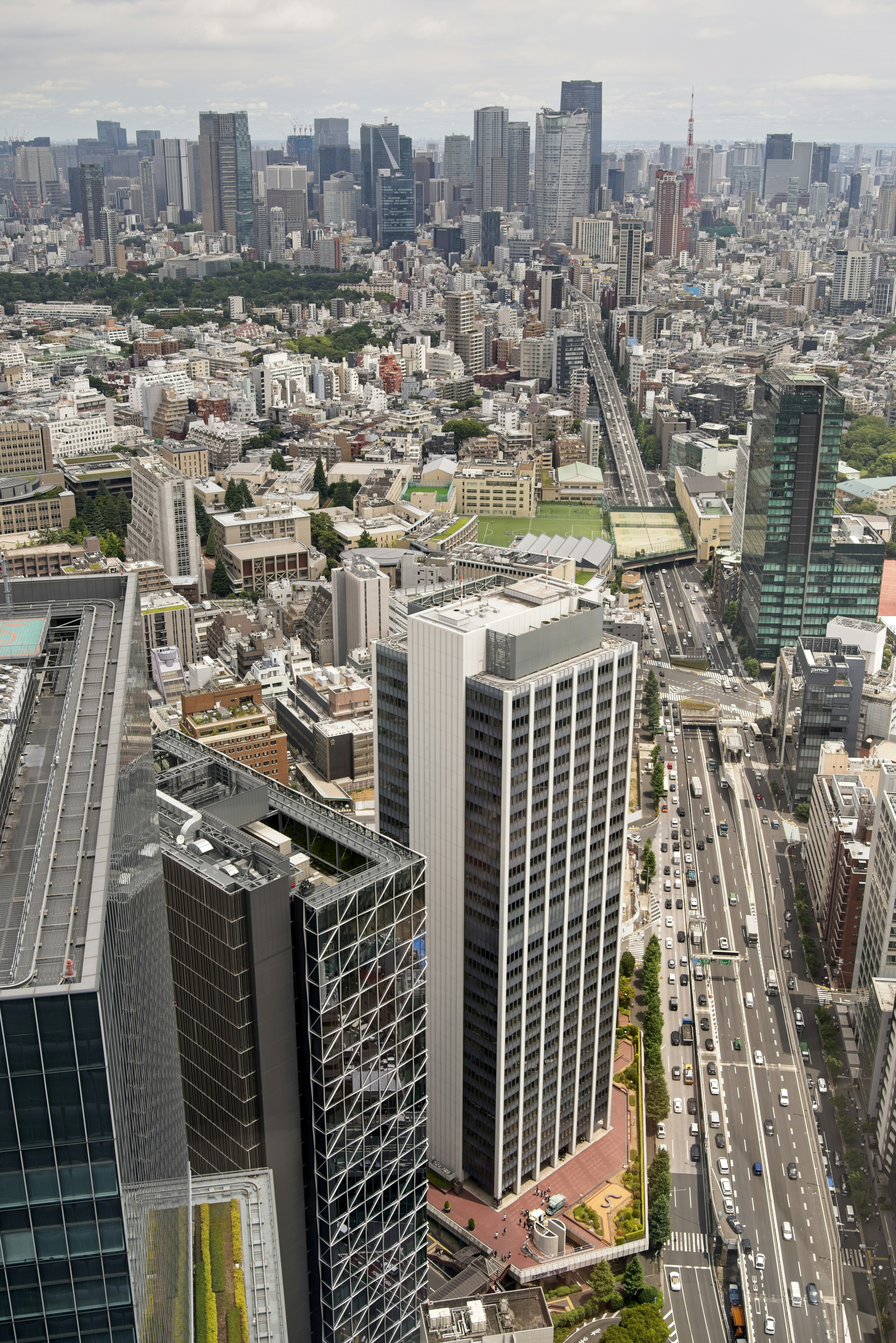 A sweeping aerial view from Shibuya Sky reveals Tokyo's dense urban sprawl stretching toward the horizon, with the distinctive red-and-white Tokyo Tower visible amid clusters of distant high-rises. A busy elevated expressway cuts diagonally through the frame, packed with traffic, while a modern glass tower rises prominently in the foreground beside a school's sports field. The layered mix of infrastructure, architecture, and rooftops captures the sheer scale of Tokyo's cityscape under a hazy, overcast July sky.