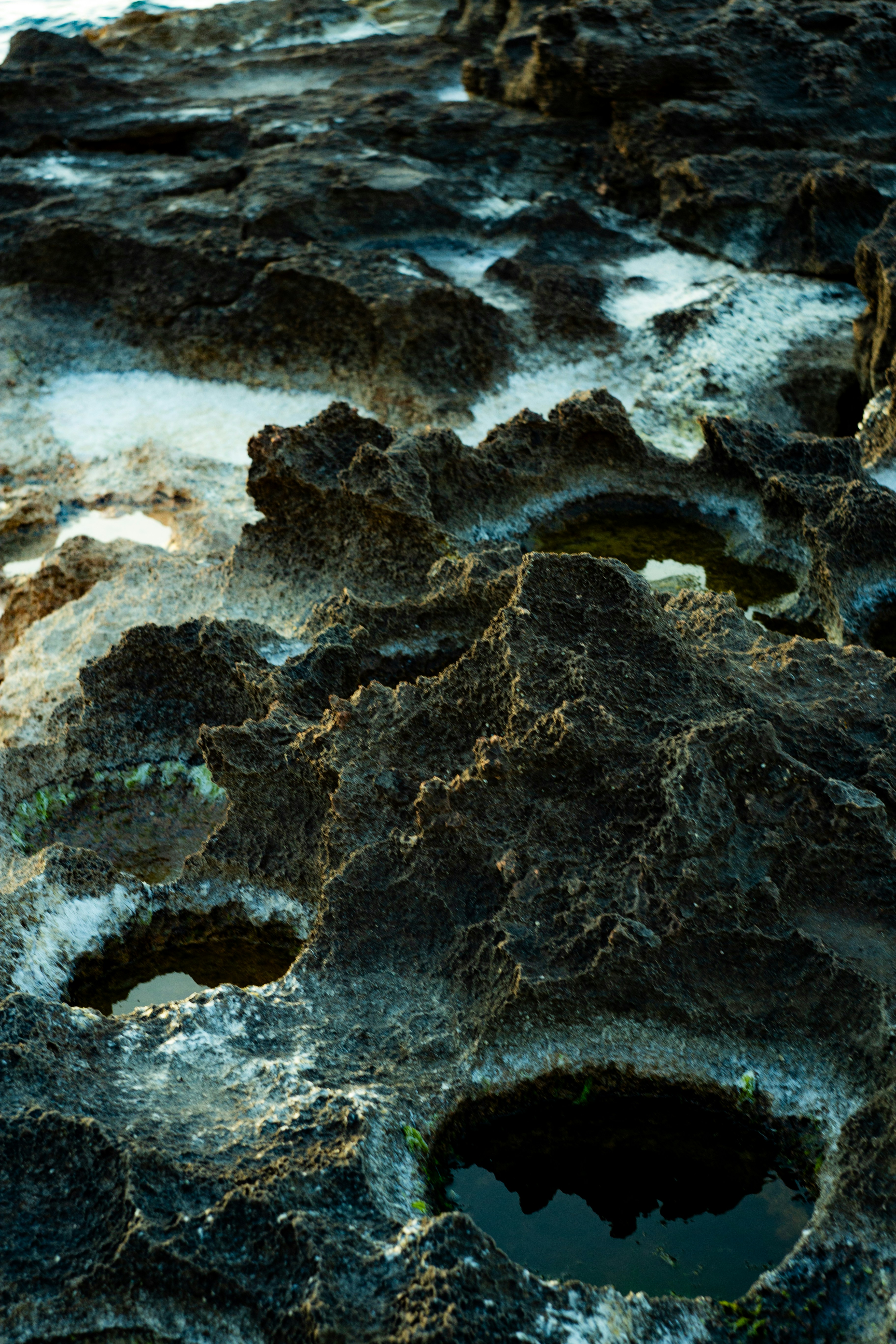 Rugged, lava-like rock formations with dark cavities and turquoise water glimpses along the coastline at low light.