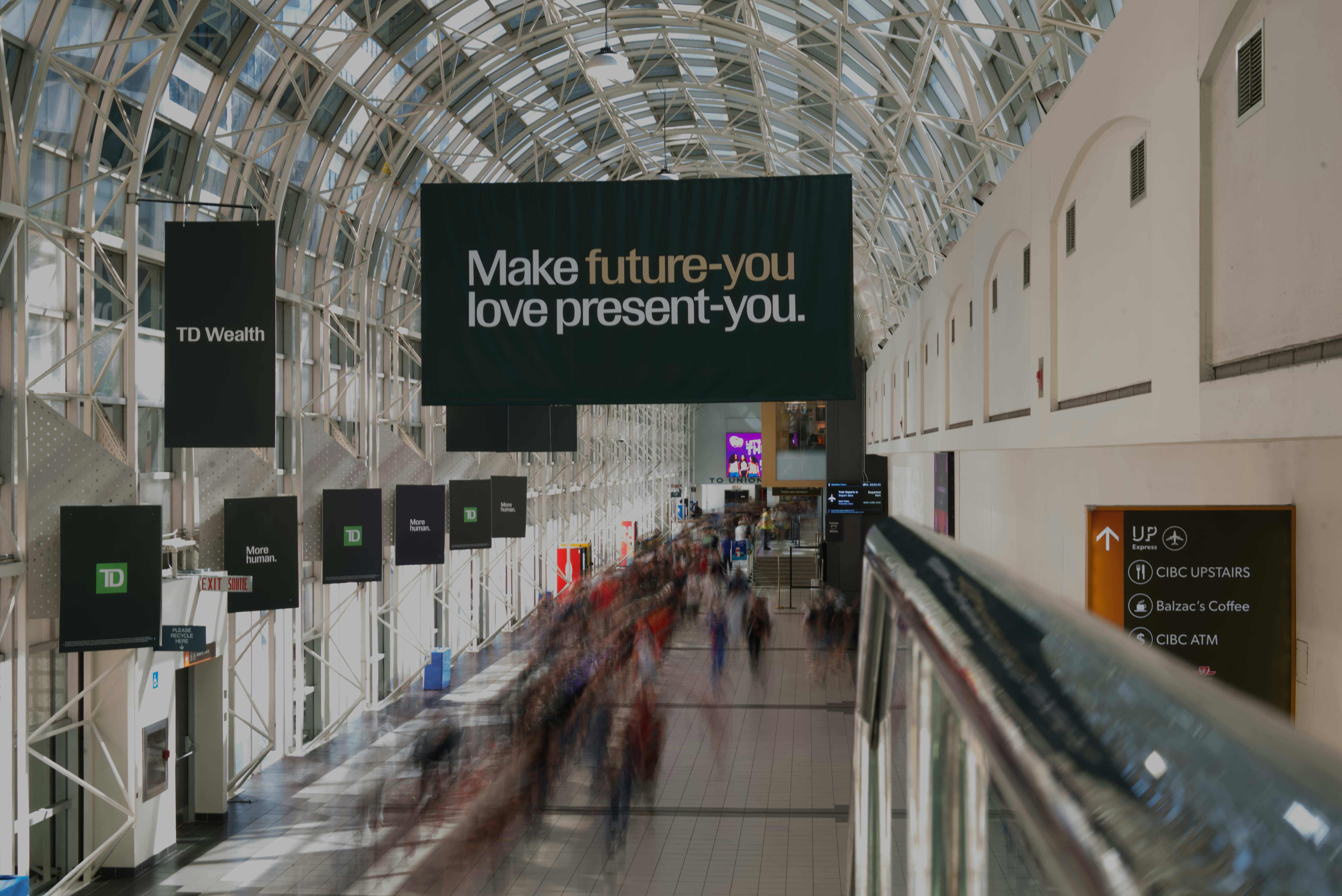 Busy airport-like terminal with a long glass ceiling; blurred figures suggest motion while a central banner reads Make future‑you love present‑you.