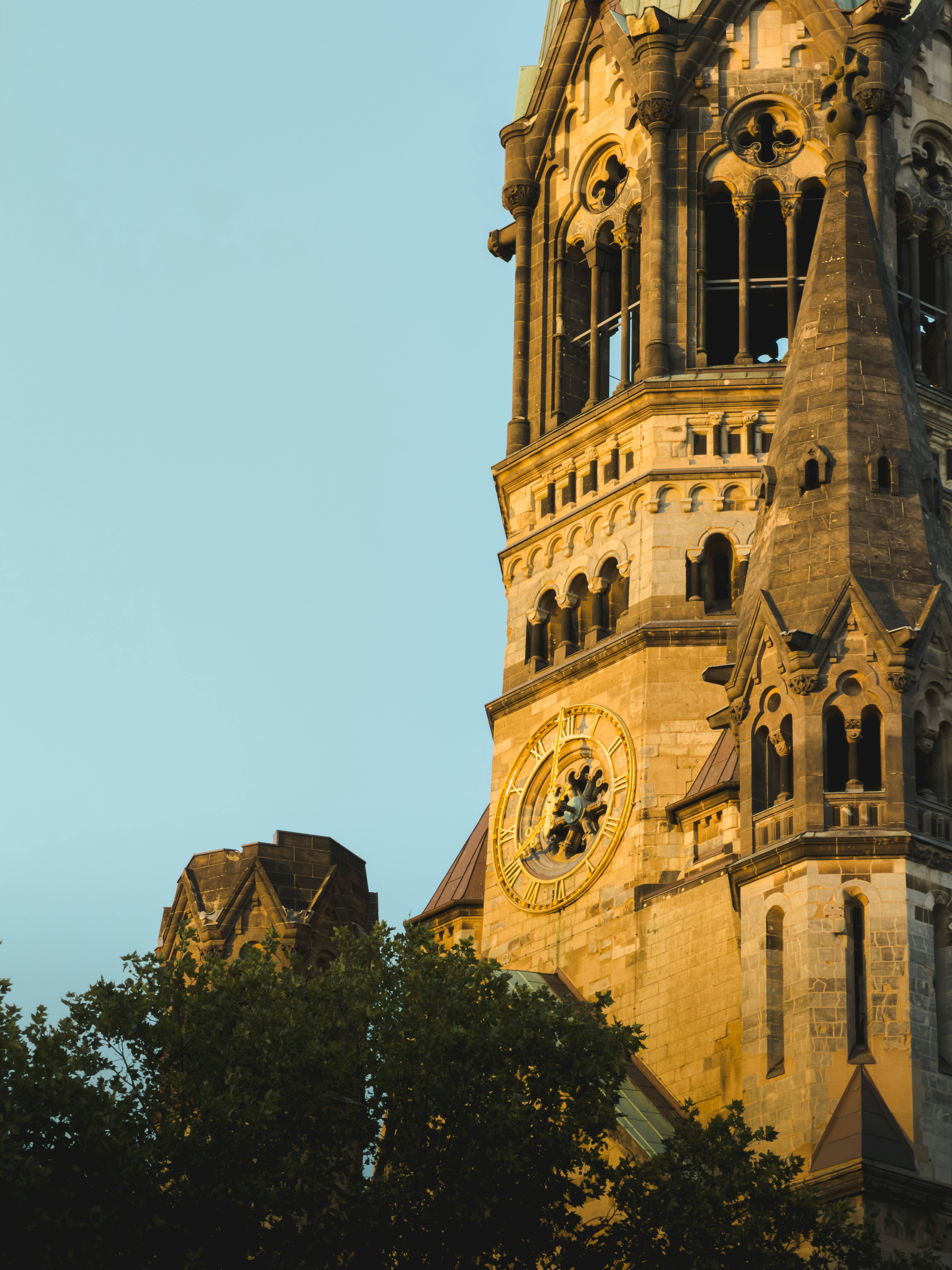 Renaissance-era clock tower rises with ornate stonework against a clear sky, its upper spire bathed in warm light and foreground trees partially framing the base.